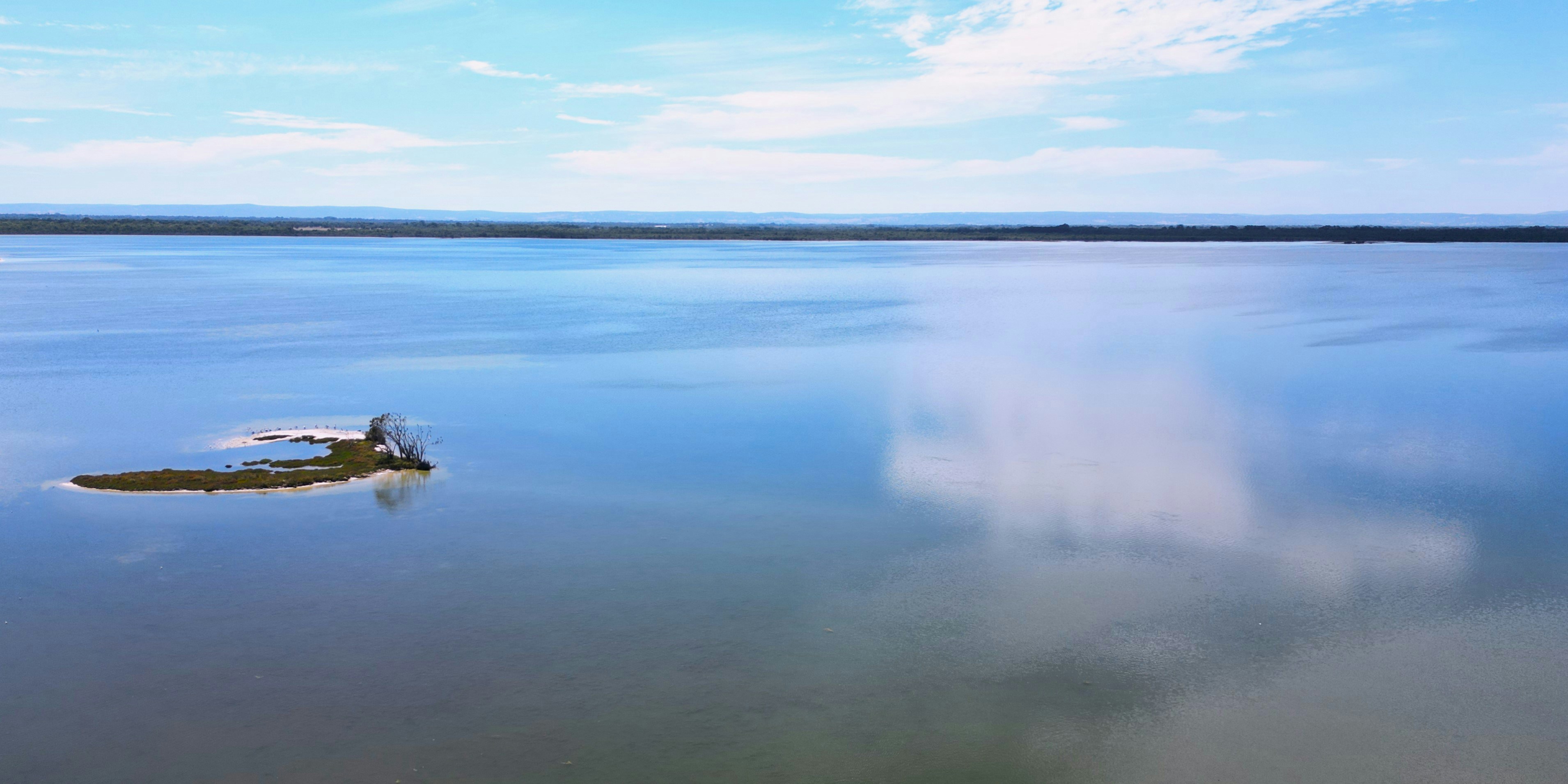 Tiny island with sparse vegetation surrounded by vast, calm lake under a clear blue sky.