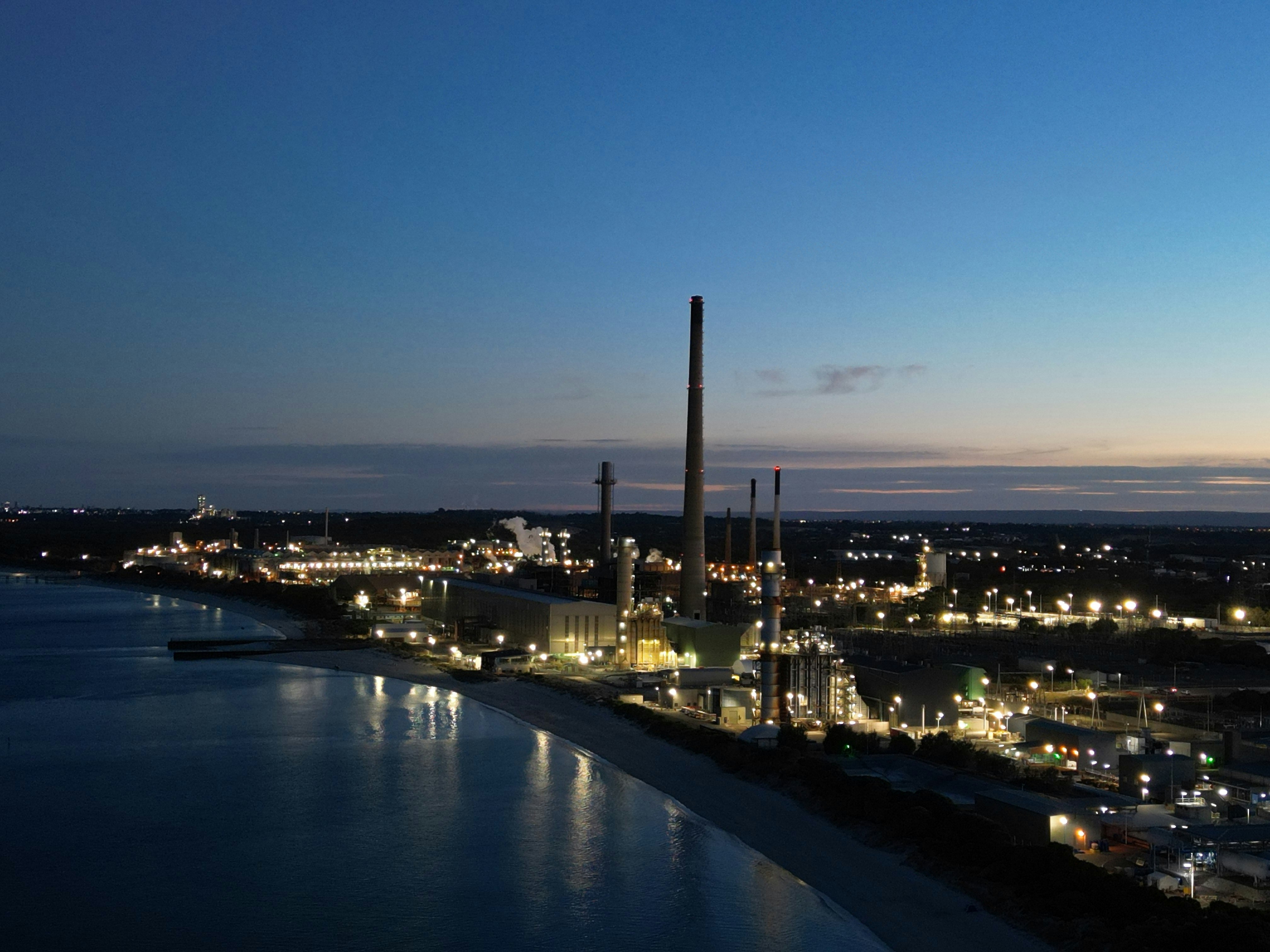 Factories lining a riverbank under a twilight sky, with lights reflecting on calm water.