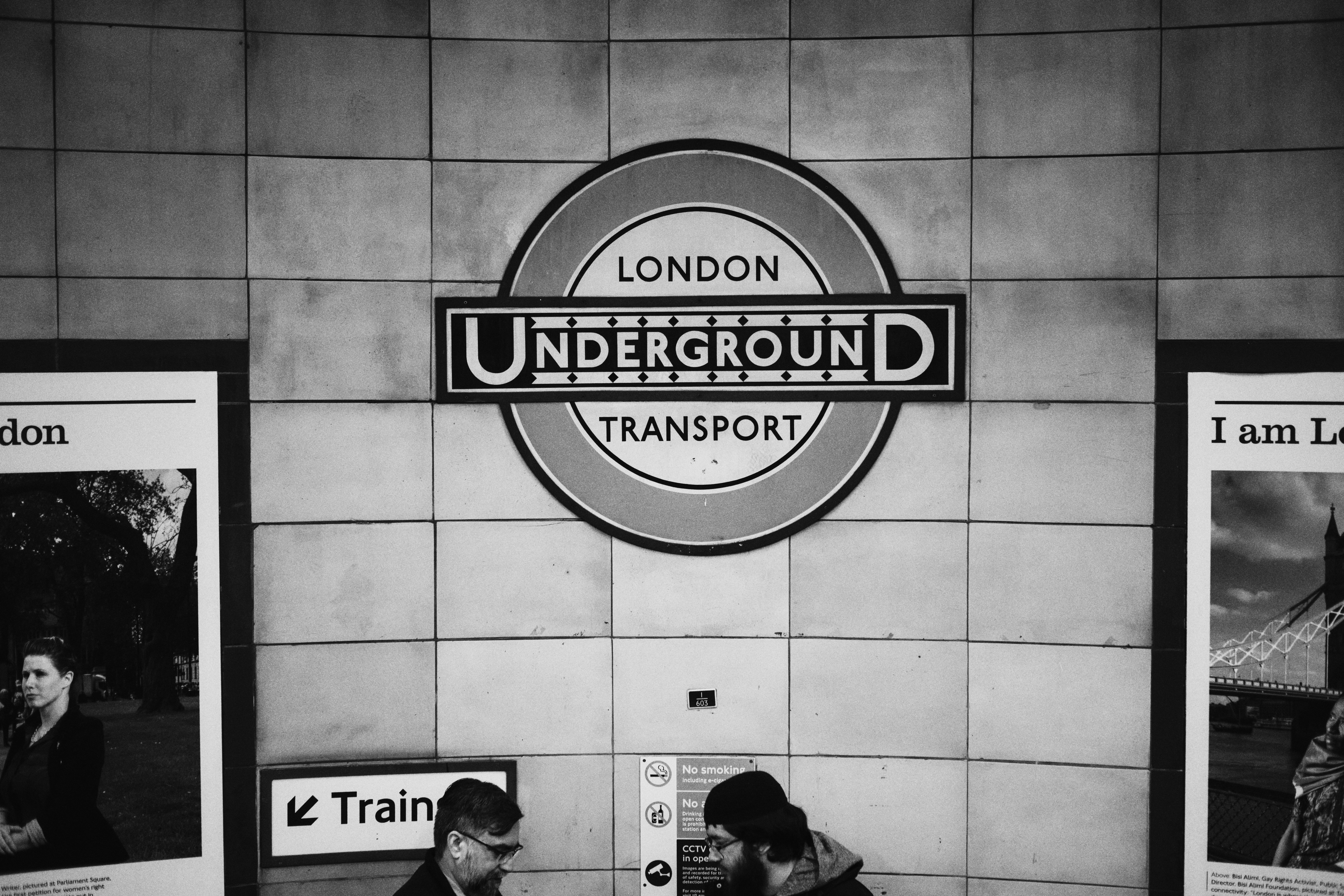 Iconic London Underground logo displayed prominently at a subway station, flanked by posters of commuters.
