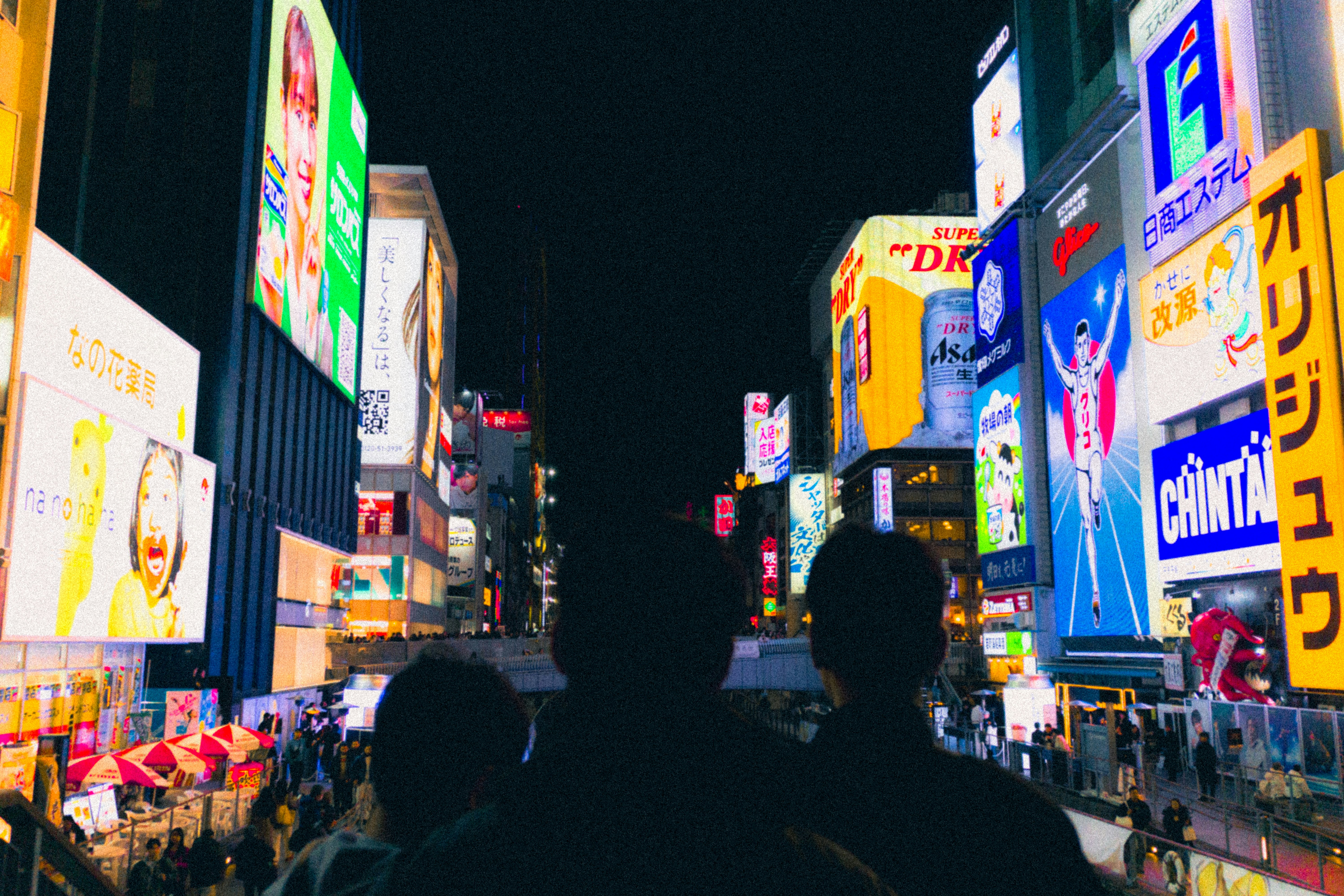 Silhouettes of three people facing vibrant neon signs in a bustling cityscape at night.