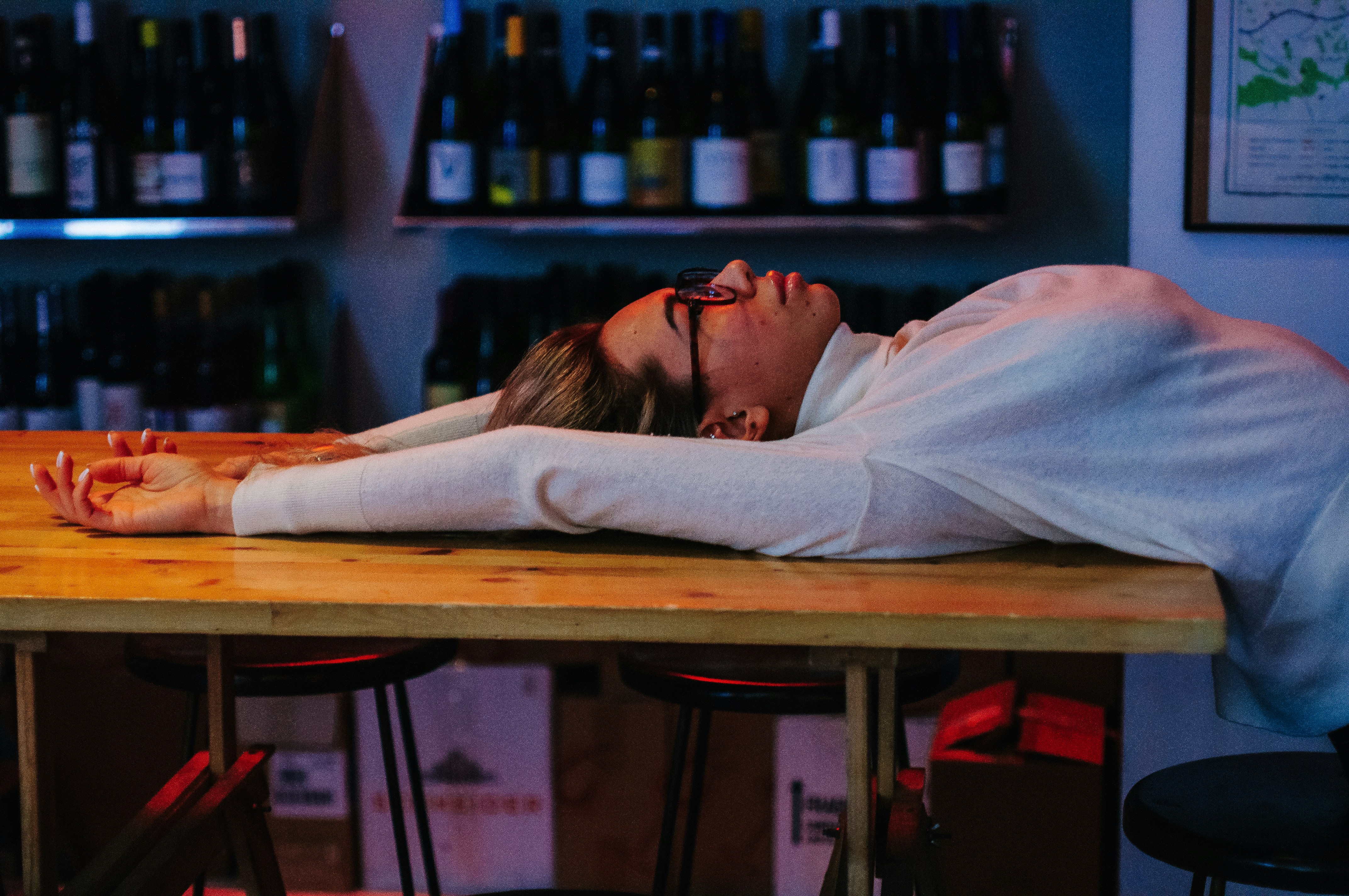 Person lying on a wooden table in a dimly lit bar with bottles on shelves in the background.