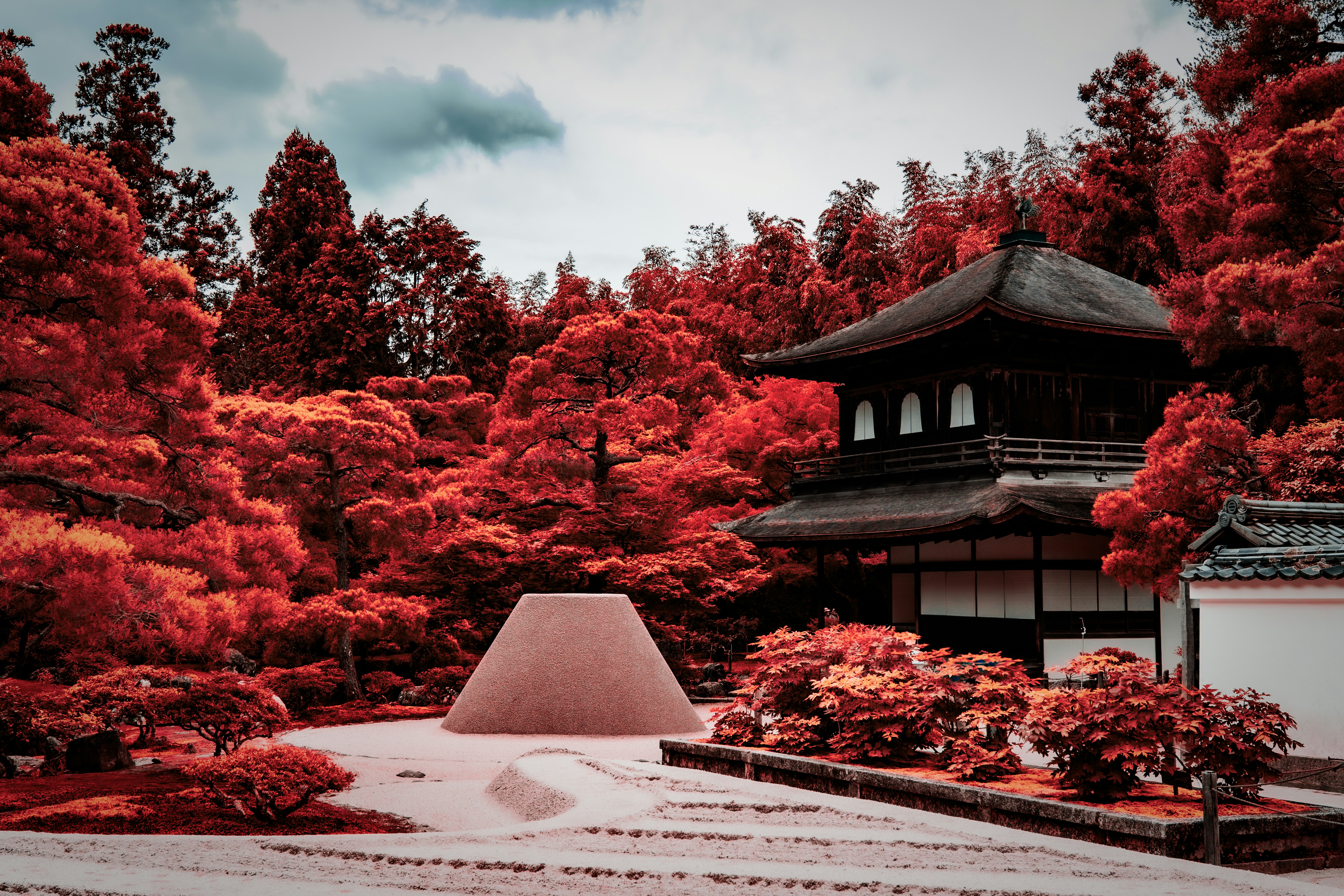 Traditional Japanese Zen garden surrounded by vibrant red foliage and a dark-roofed building under a cloudy sky.