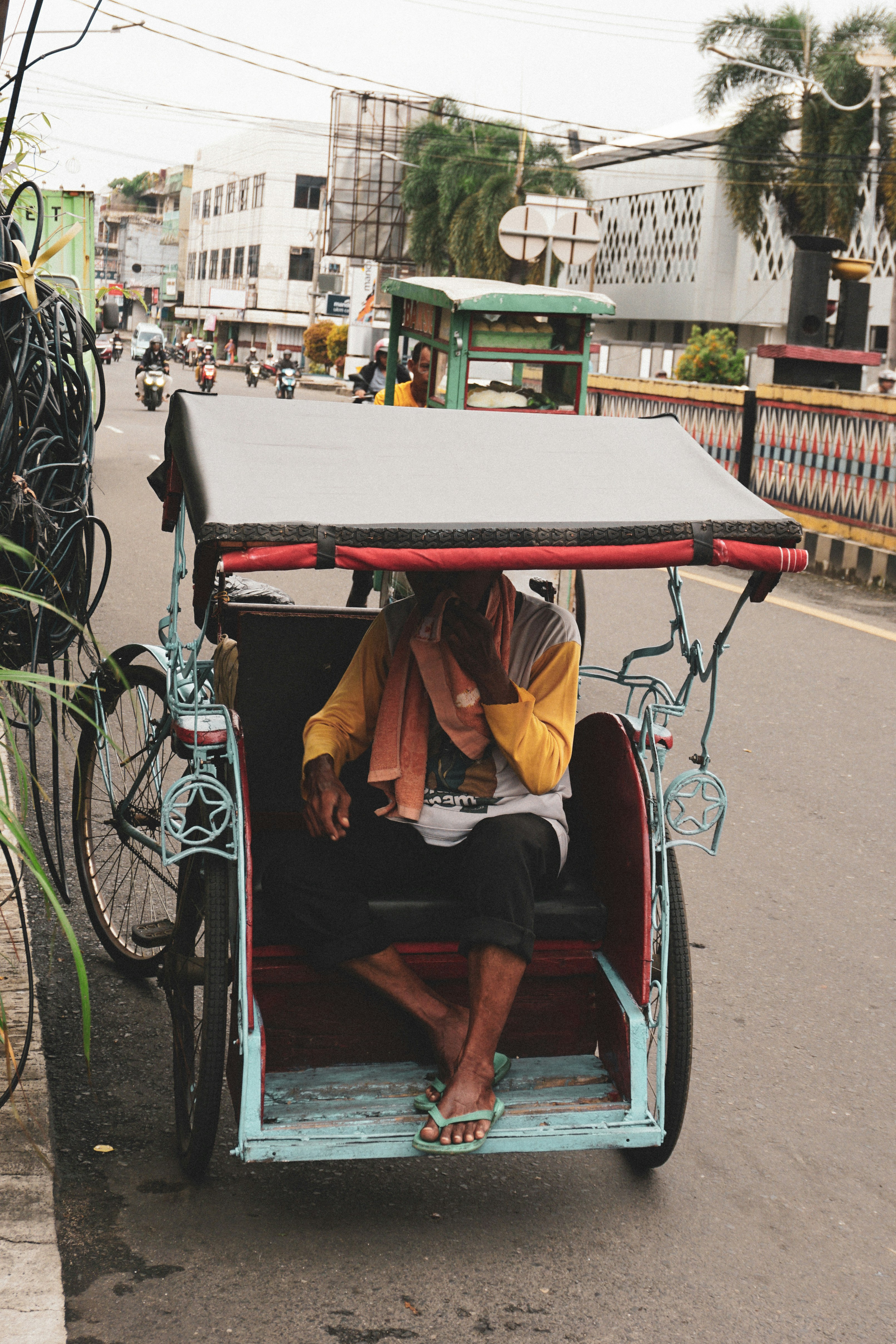 An elderly man resting in a cycle rickshaw, partially obscured by his hand, amidst a bustling street scene filled with vehicles and greenery.
