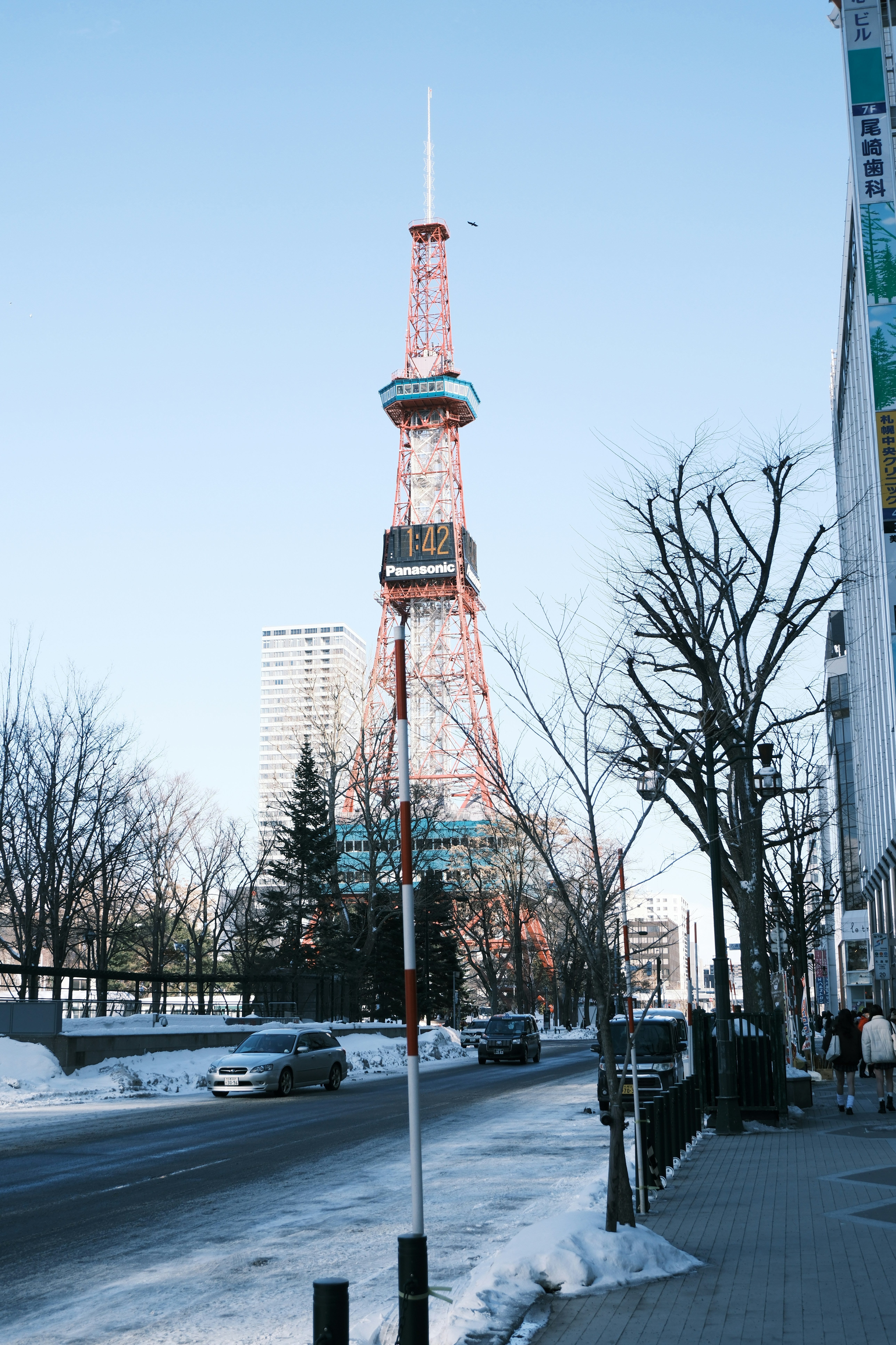 Photograph of a red lattice tower with a digital clock, rising above a snow-covered city street framed by bare trees.