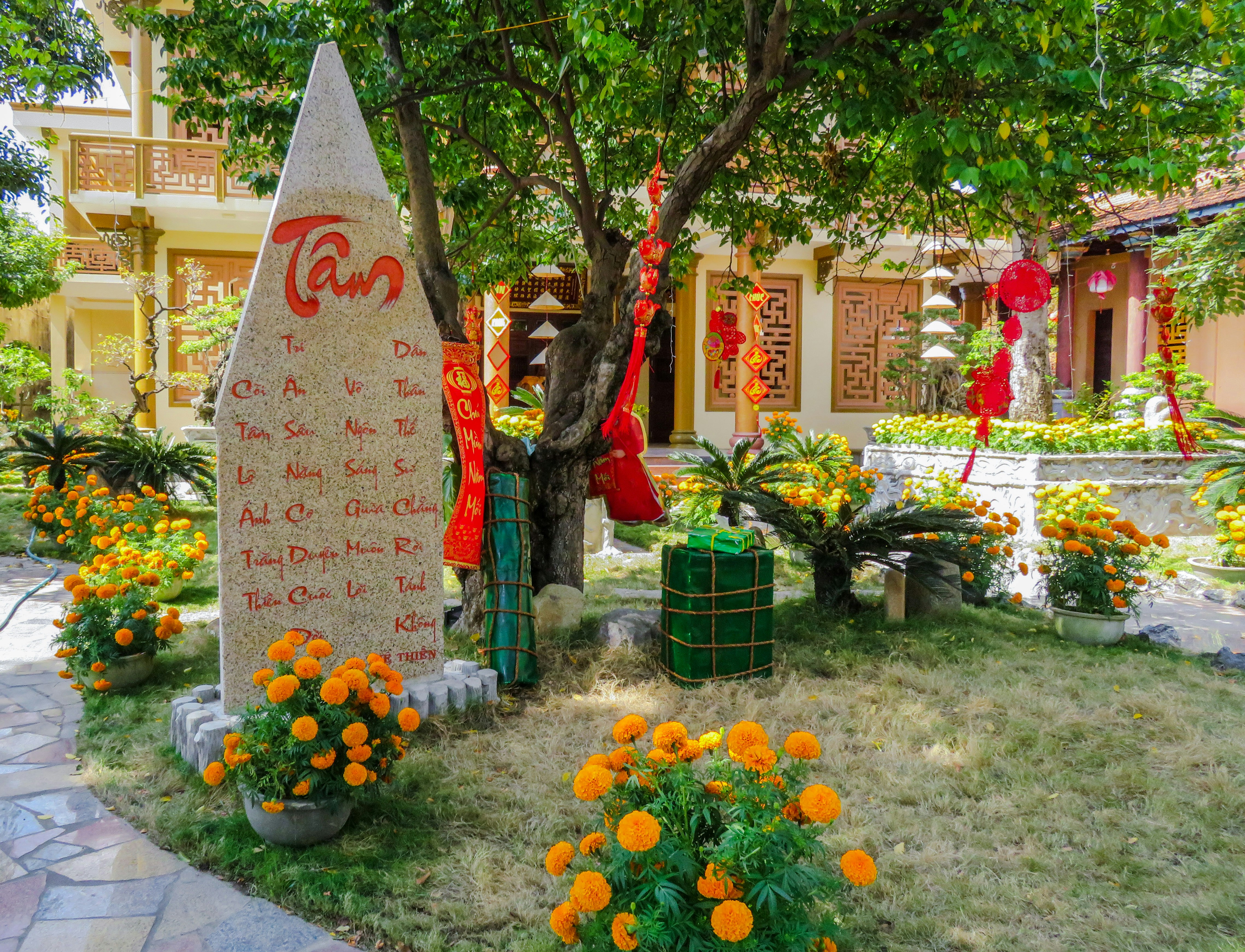 Decorative stone with Vietnamese script surrounded by marigolds and festive red ornaments under a tree.