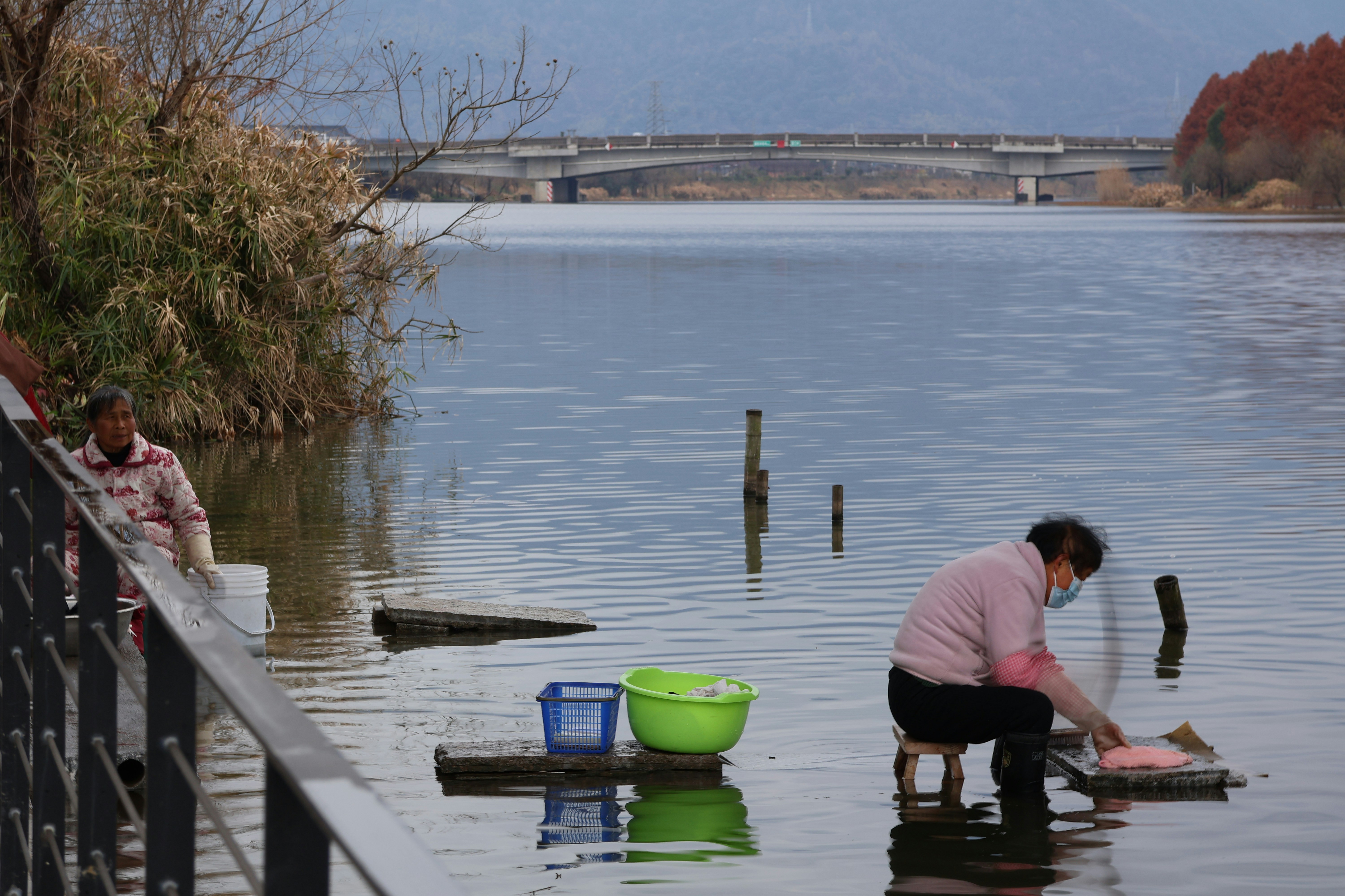 Two people wash clothes in a calm river, surrounded by autumn foliage and distant mountains.