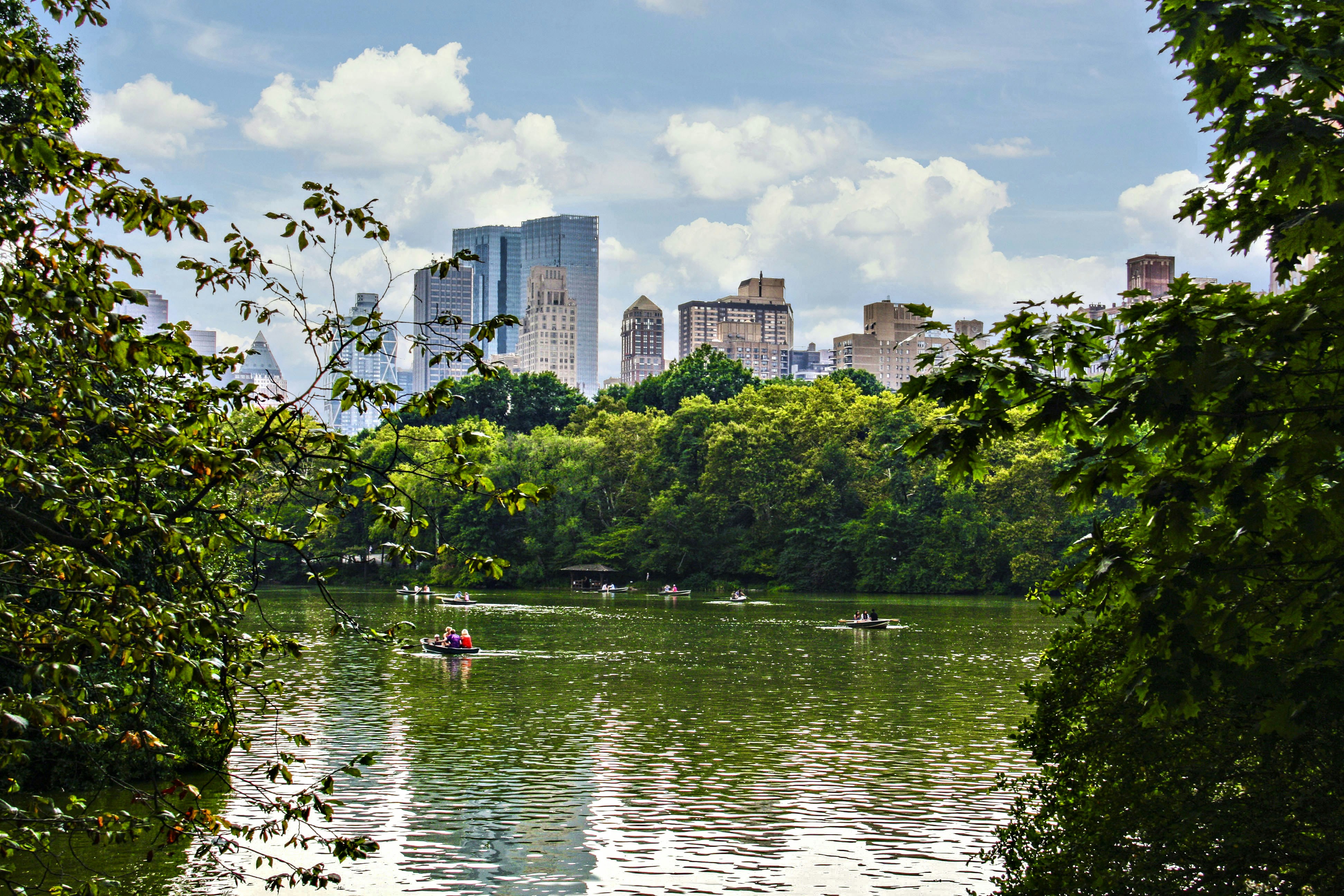City skyscrapers rise above lush greenery and a tranquil lake with boats under a partly cloudy sky.