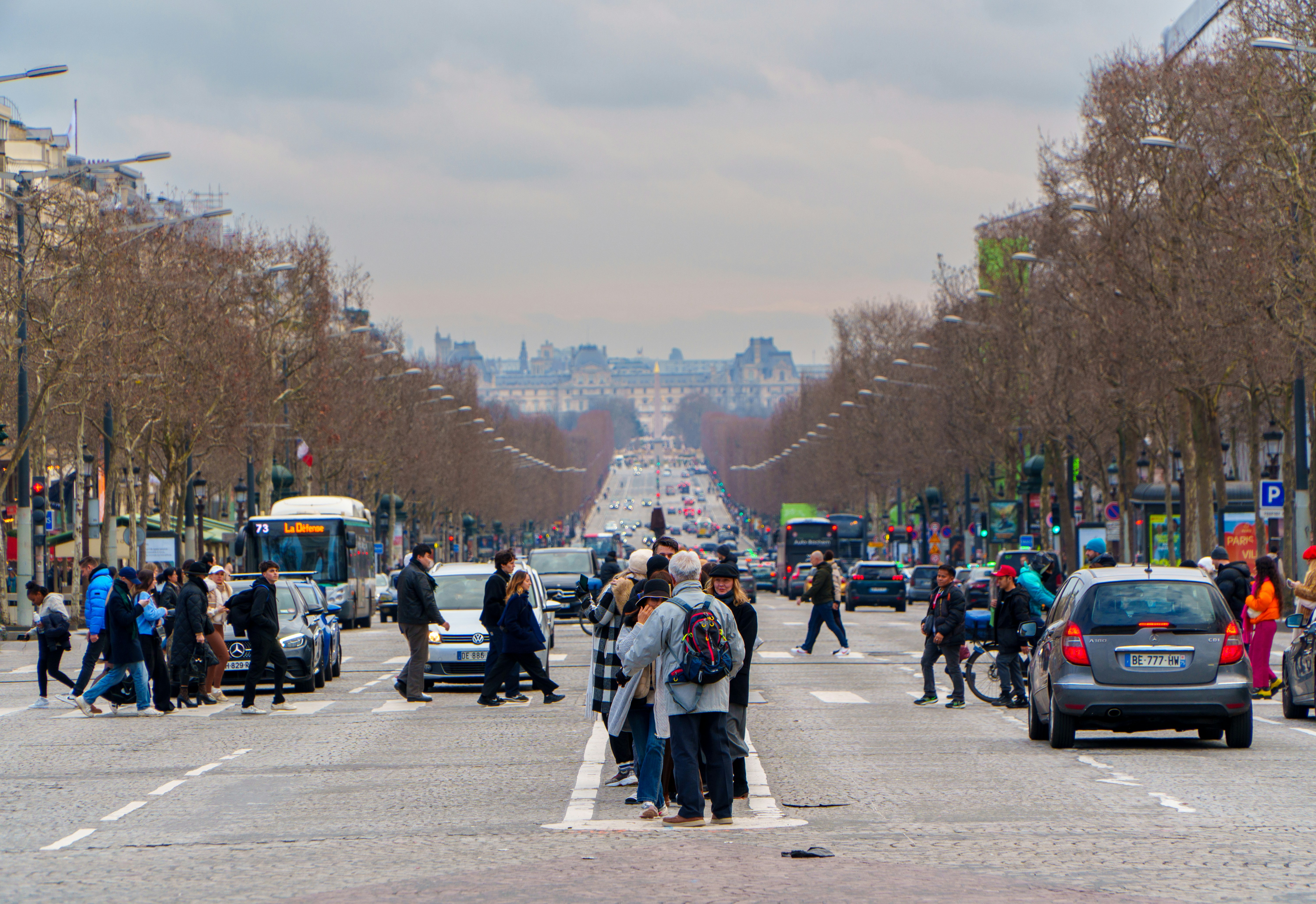 People crossing a busy Parisian avenue with the Arc de Triomphe in the distance.