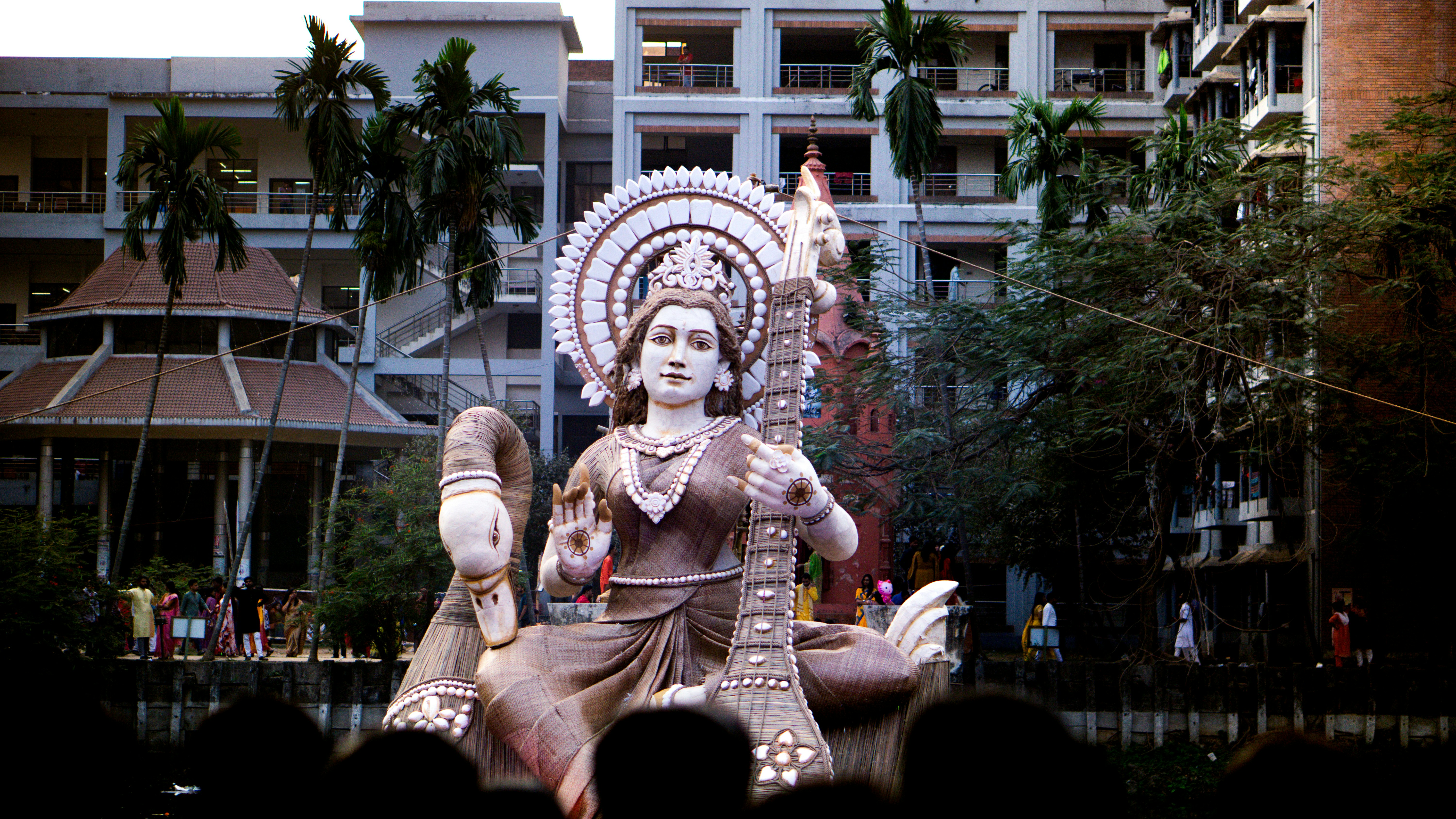 Statue of a seated deity playing a musical instrument with a swan, set against a backdrop of modern buildings and palm trees.