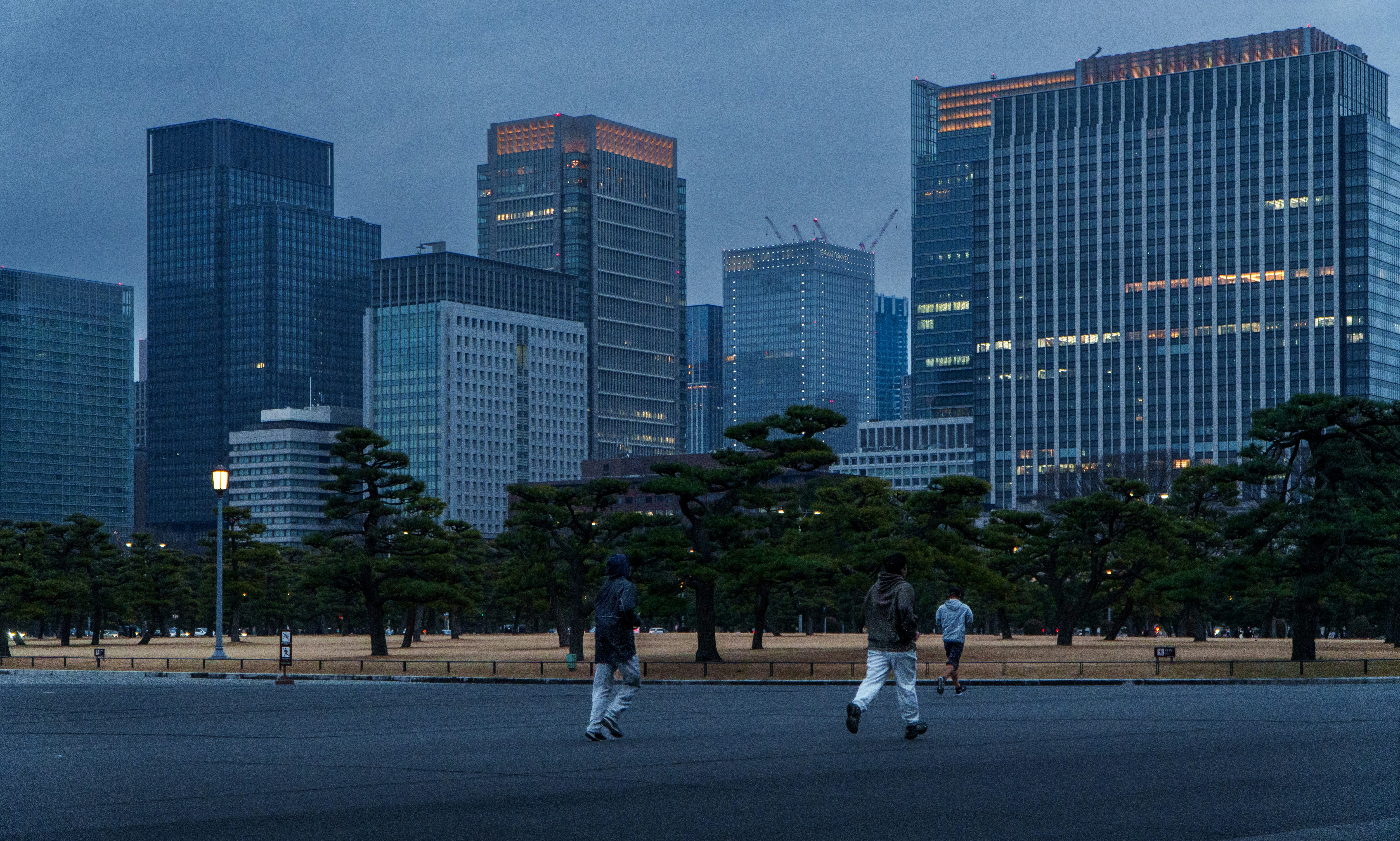 Runners move through the Imperial Palace gardens against the backdrop of Tokyo's skyscrapers under a dim sky.