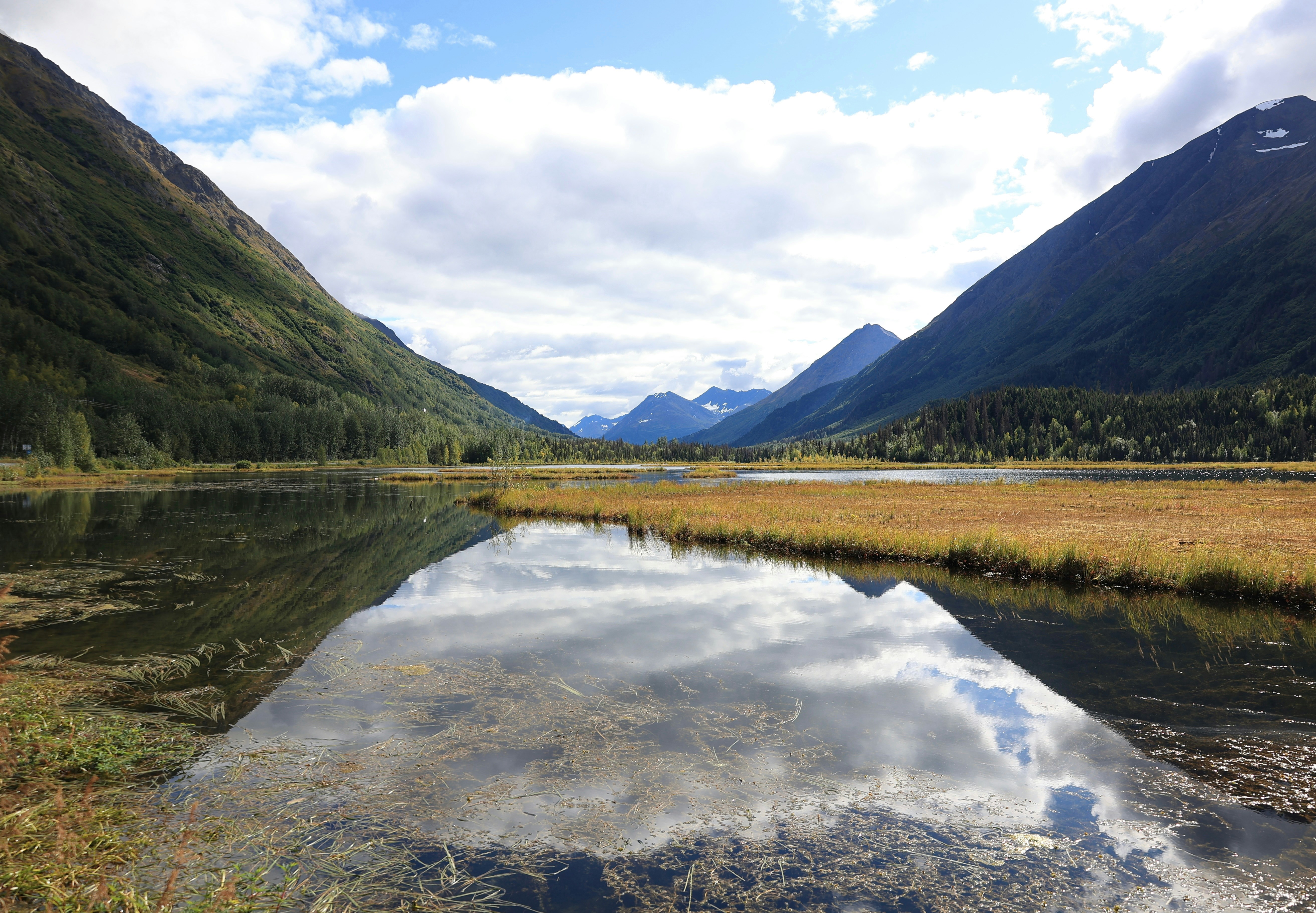 Clouds and mountains reflect in a tranquil lake surrounded by lush greenery.