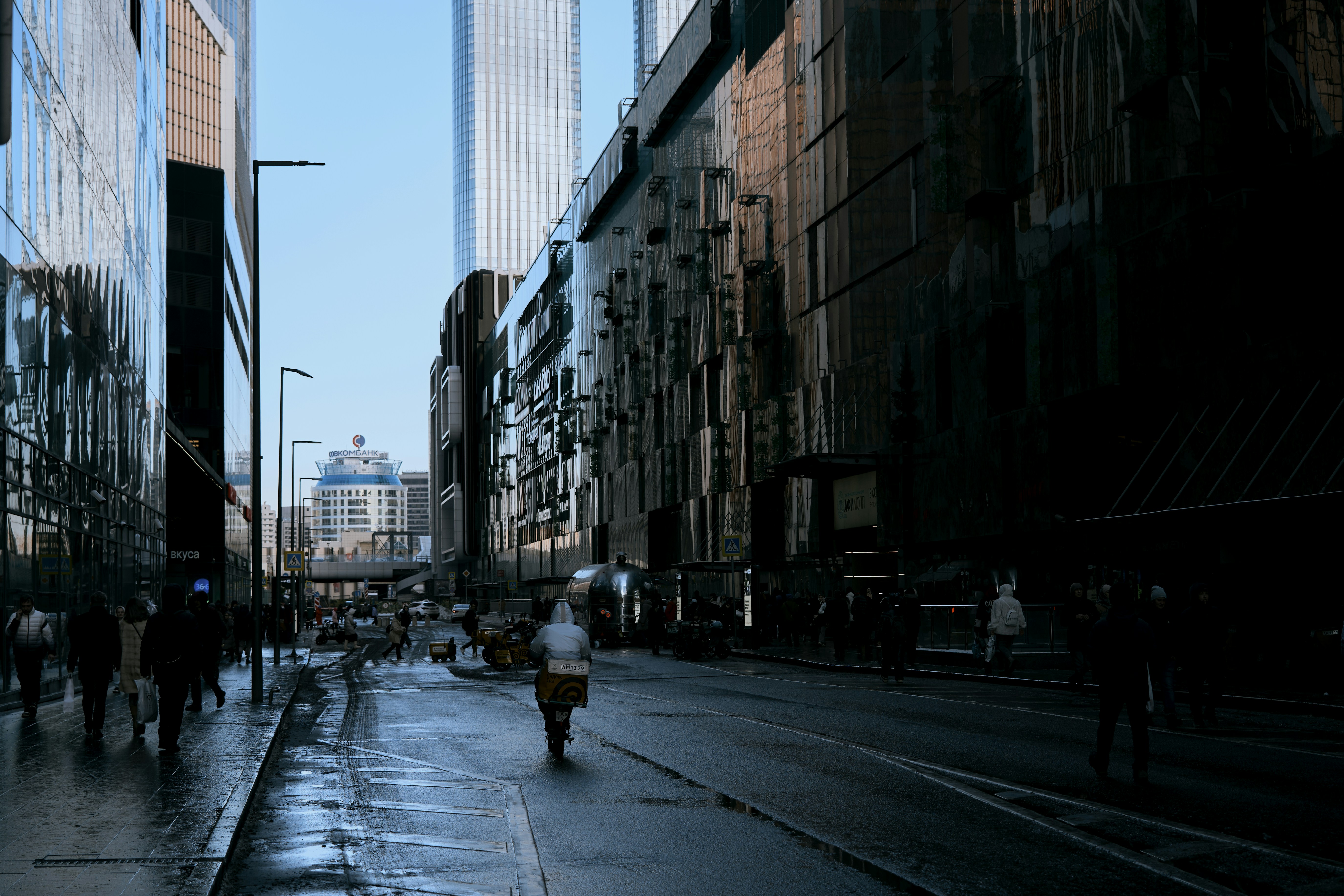 City street with reflective skyscrapers flanking a busy road, where a cyclist navigates through pedestrians.
