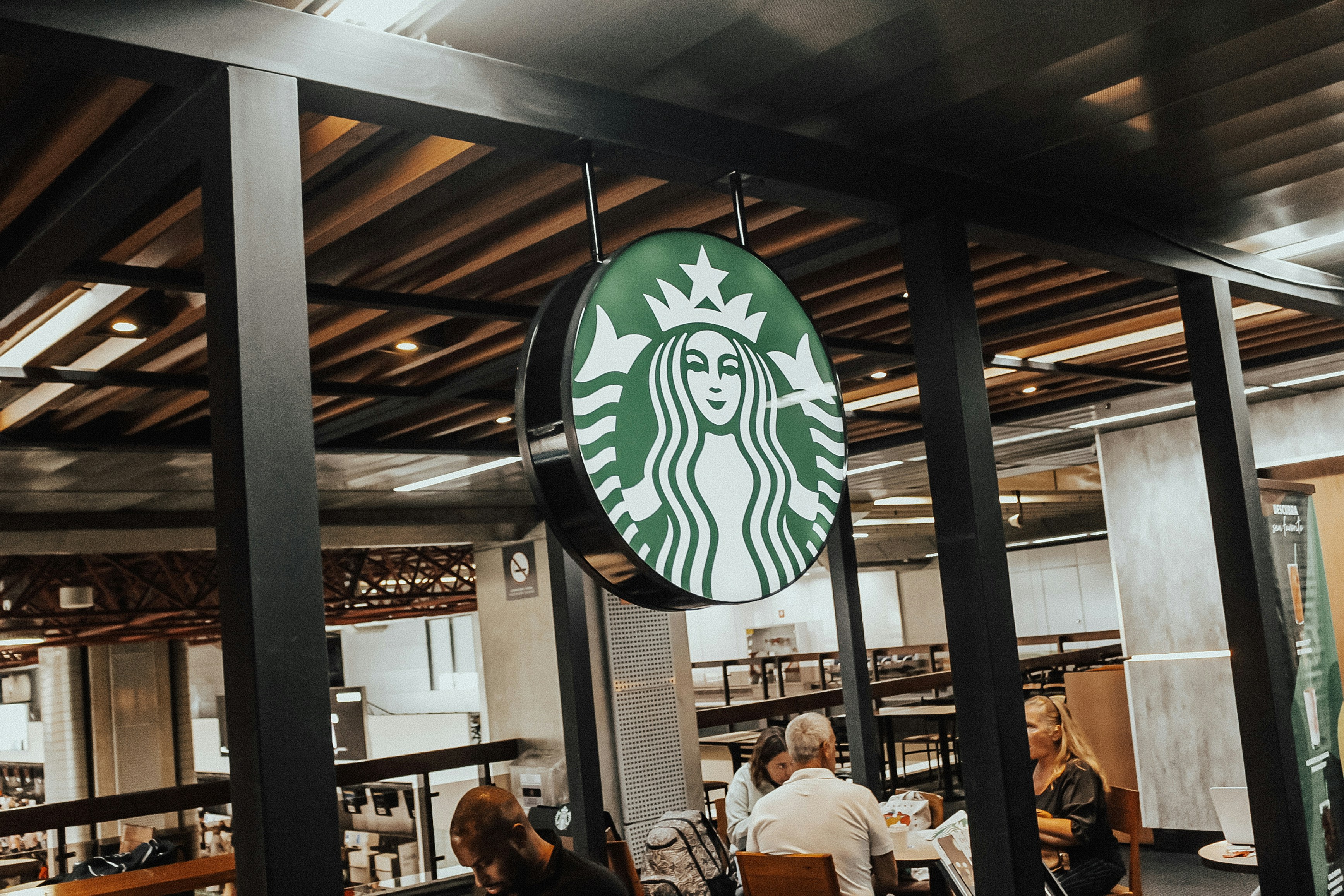 Starbucks sign hanging inside a modern airport café with people seated at tables.