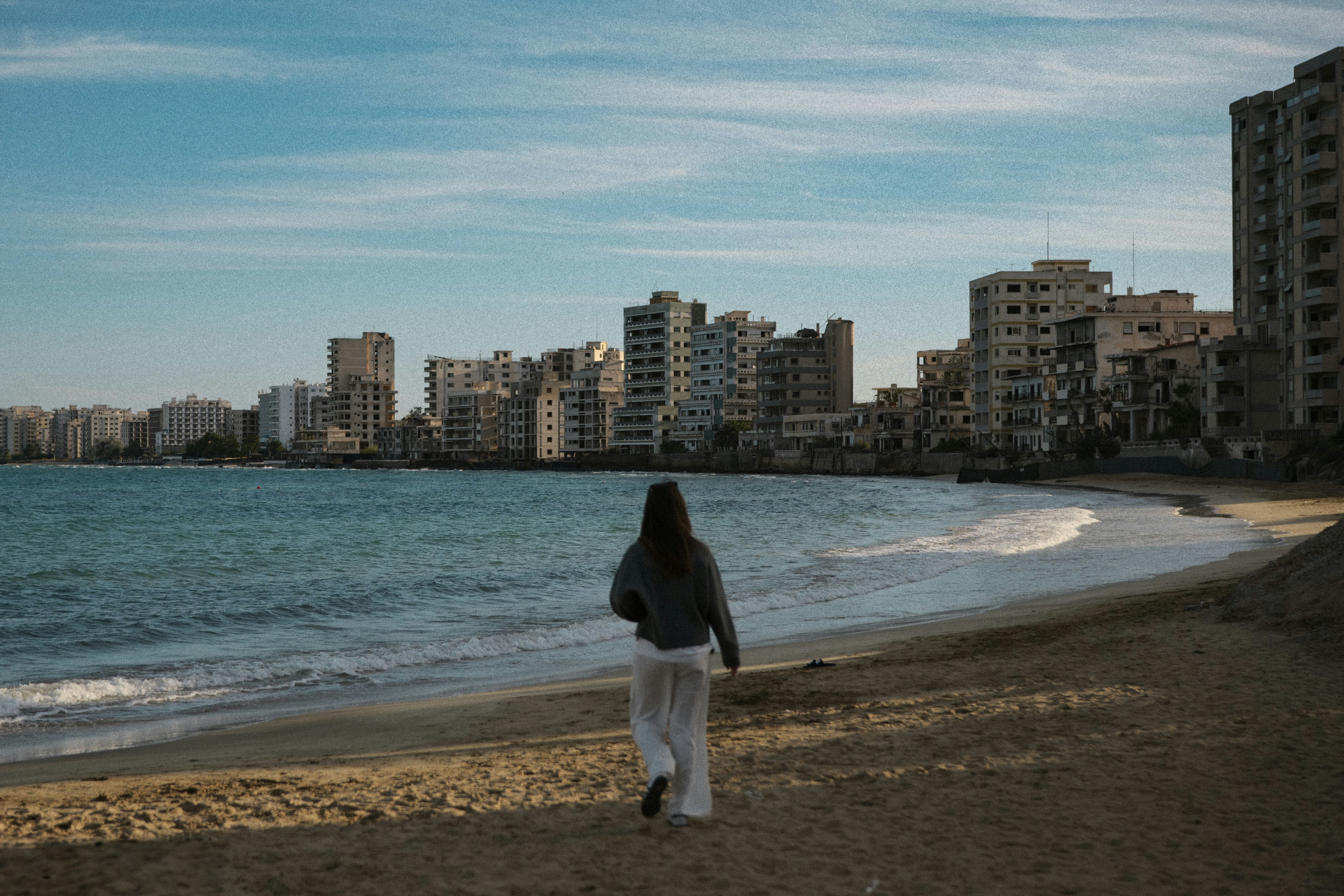Person walking along a deserted beach with a backdrop of empty high-rise buildings under a blue sky.