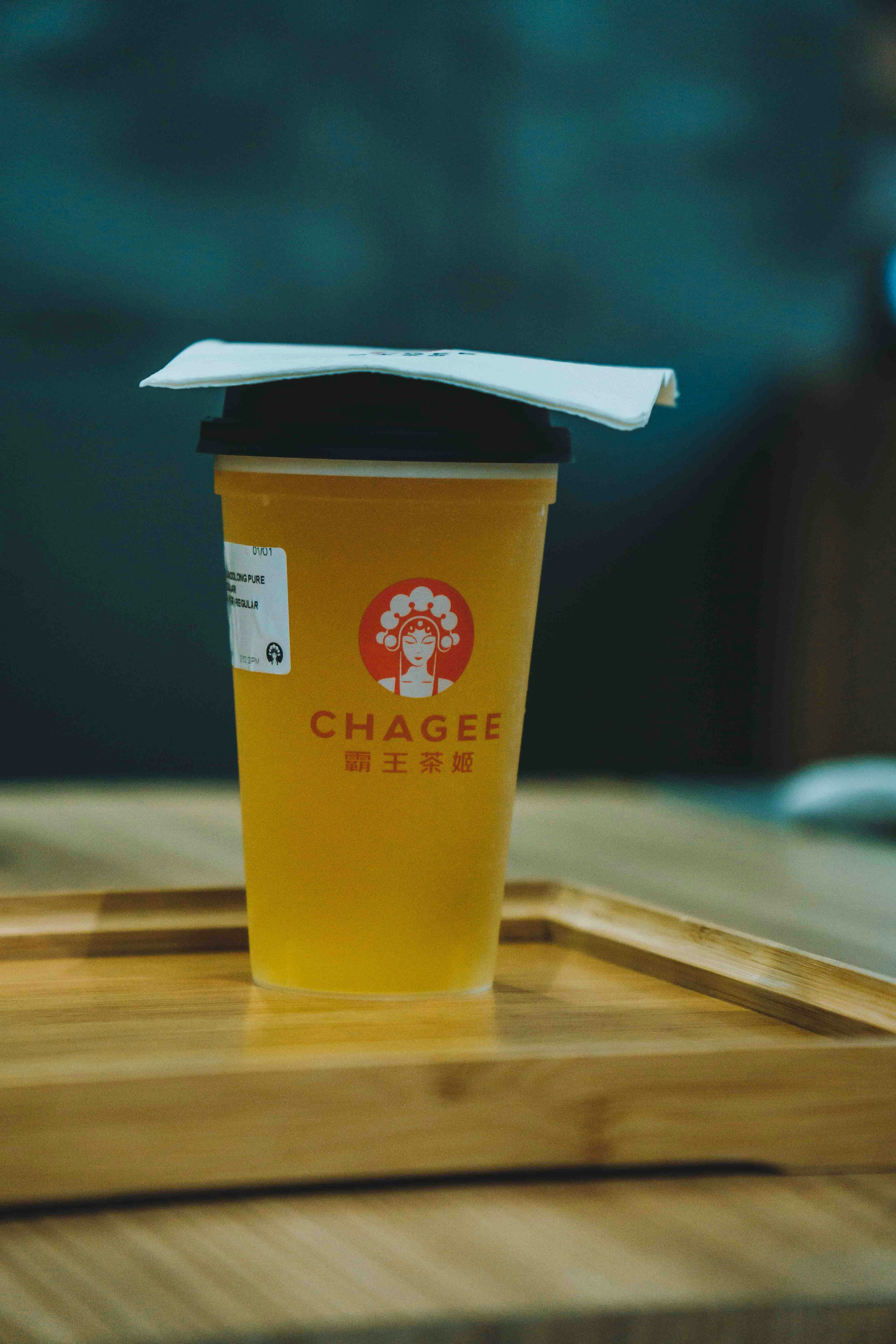 A clear beverage in a branded cup rests on a wooden tray, showcasing the Chagee logo. The background features a softly blurred interior, enhancing the focus on the drink.