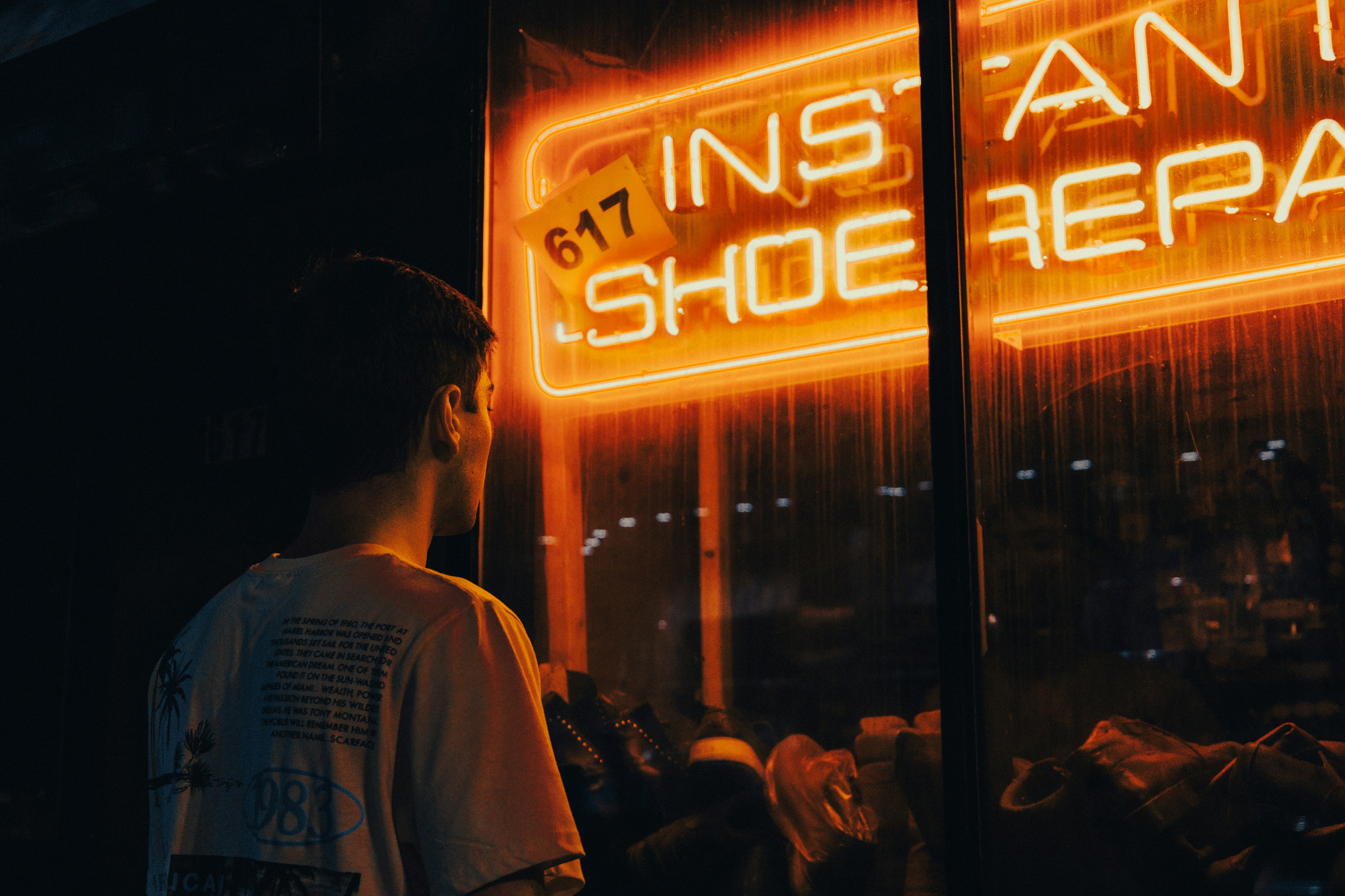 Person gazing into a shop window with a bright orange neon sign reading 'Instant Shoe Repair'.