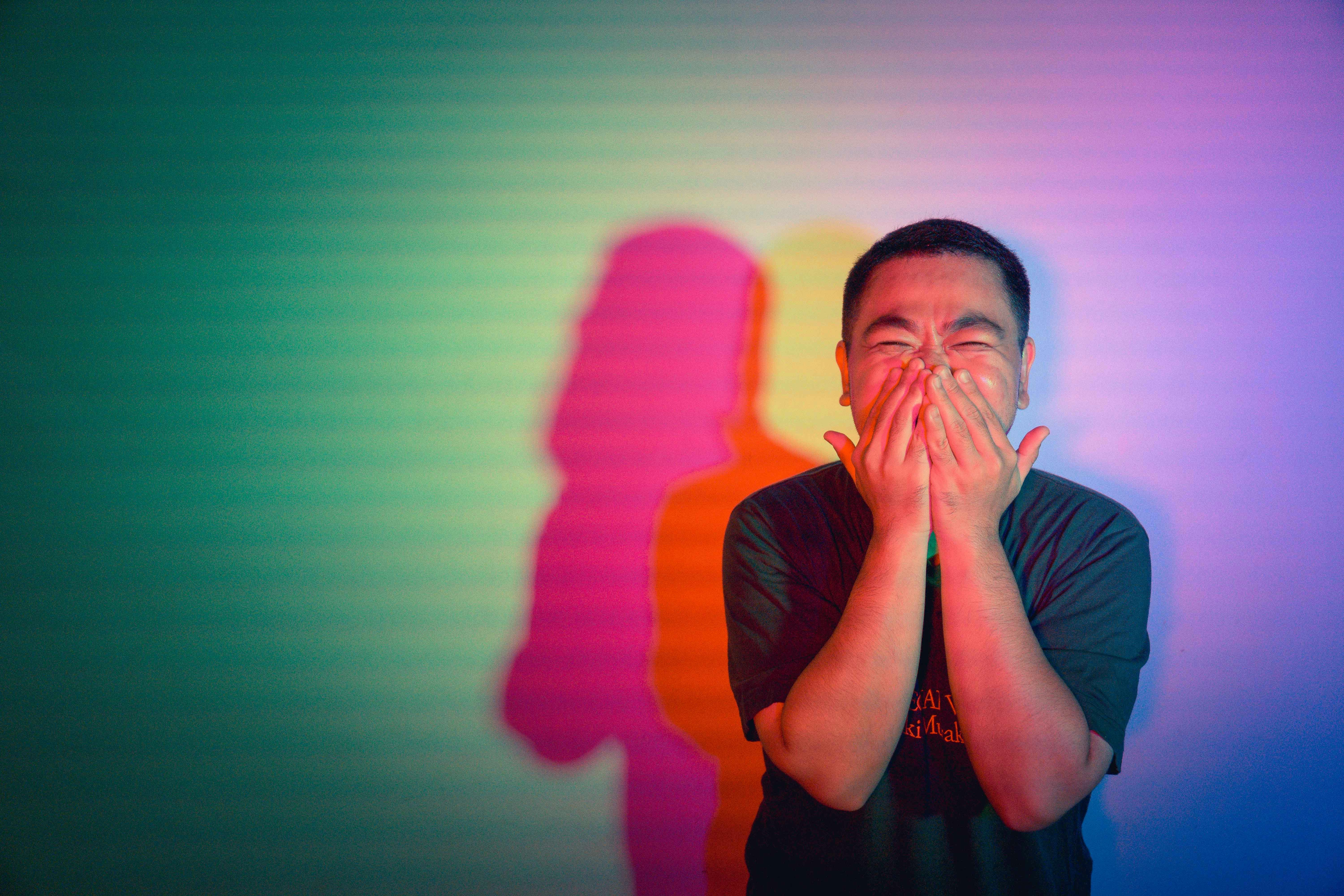 Person laughing with hands covering face, casting a multicolored shadow on a gradient wall.