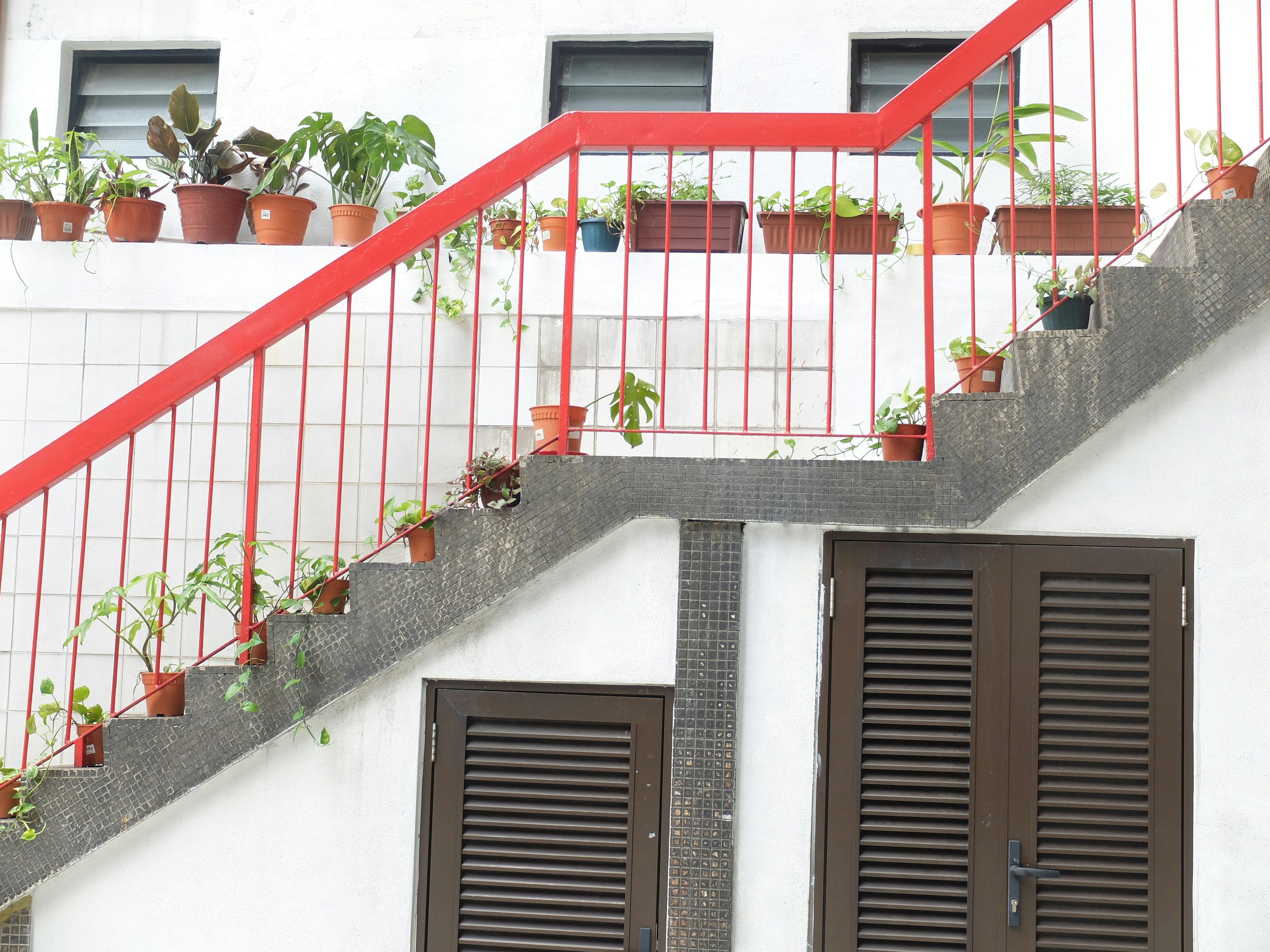 Diagonal red railing frames a stairwell along a white facade, with rows of potted plants lining the balcony above.