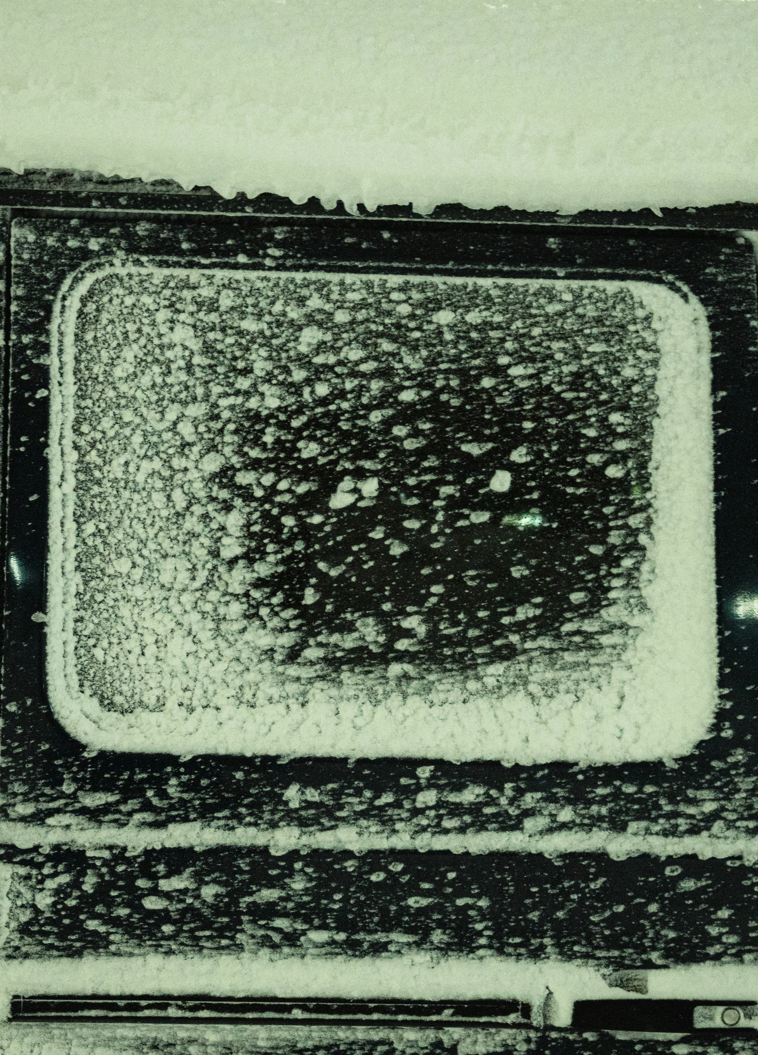 A snow-covered surface obscures a window, creating a textured pattern of frost and ice. The scene conveys the quiet stillness of a winter day.