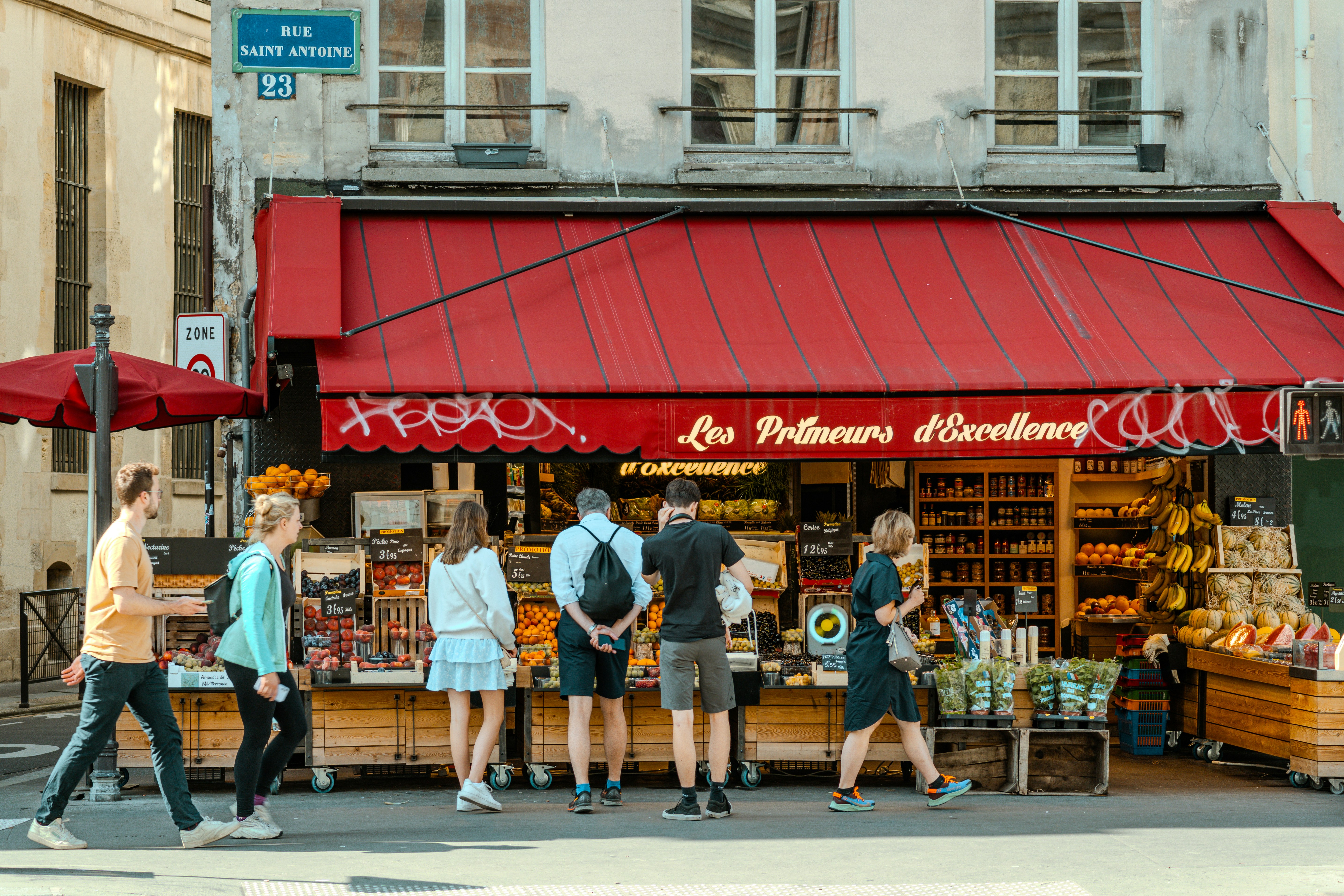 Market Stalls Under a Red Awning: A Line of Shoppers Engages Fruit and Baskets
