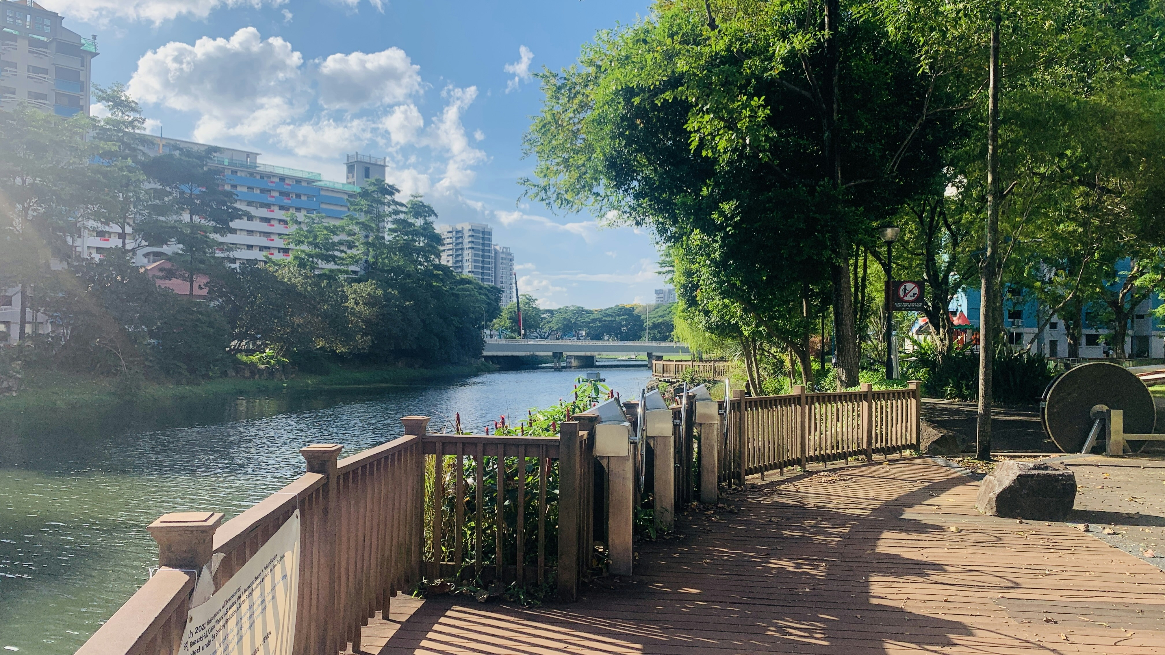 Wooden walkway beside a tranquil river under a bright blue sky with scattered clouds and urban buildings in the background.