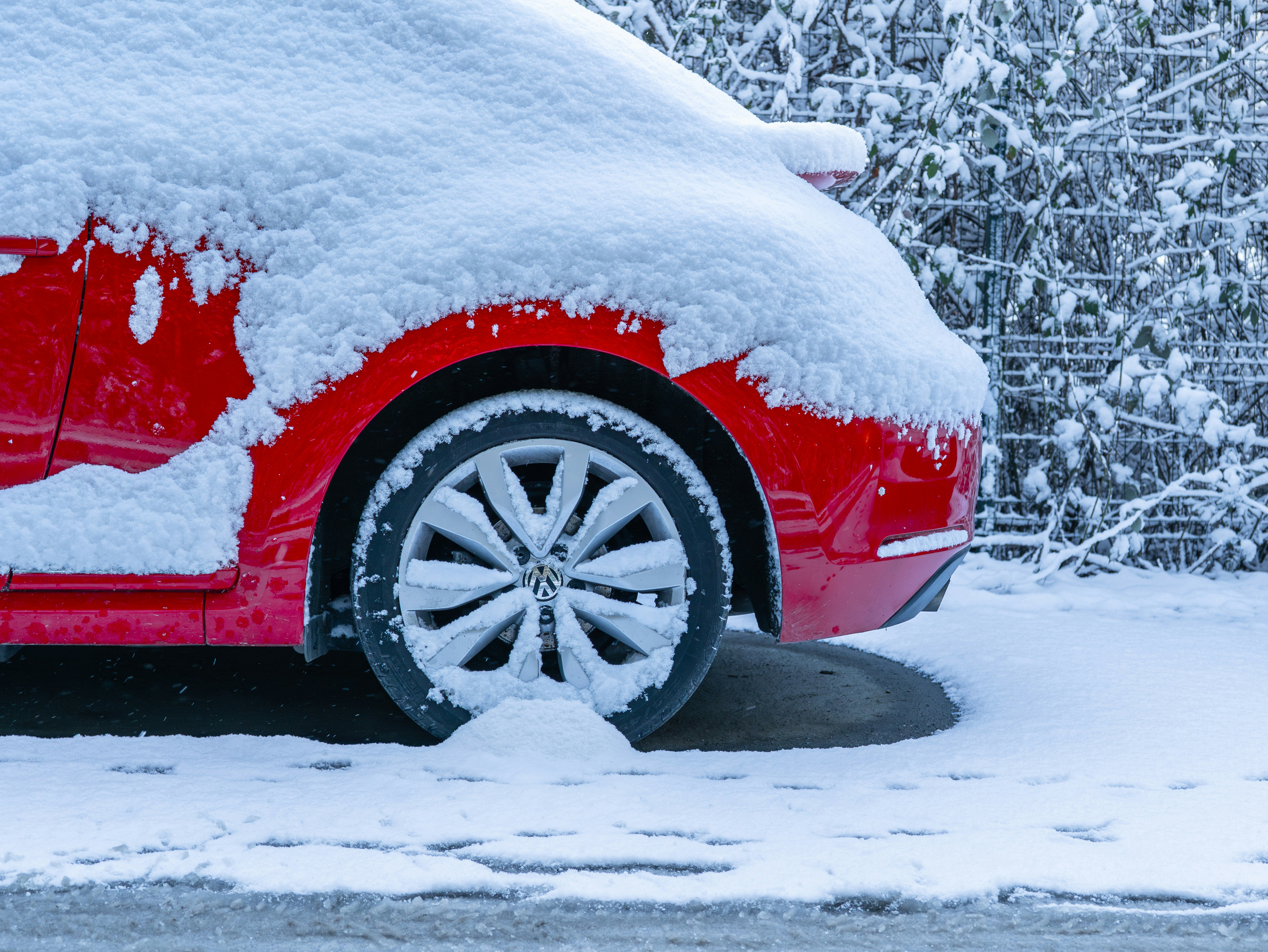 Red car partially covered in snow, parked near a snow-draped hedge on a frosty day.