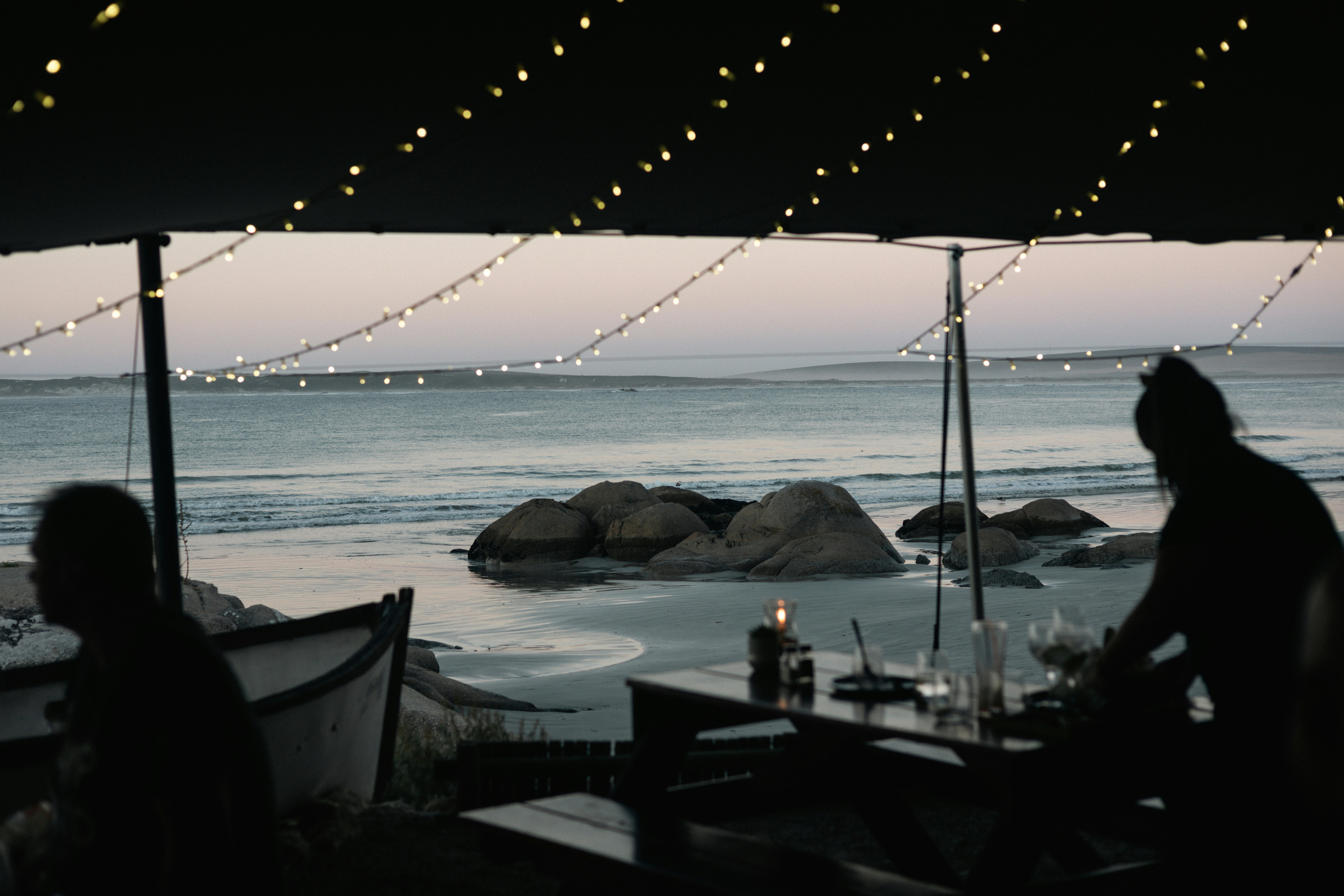 Coastal view from a café with string lights illuminating the foreground and rocks in the gentle surf.