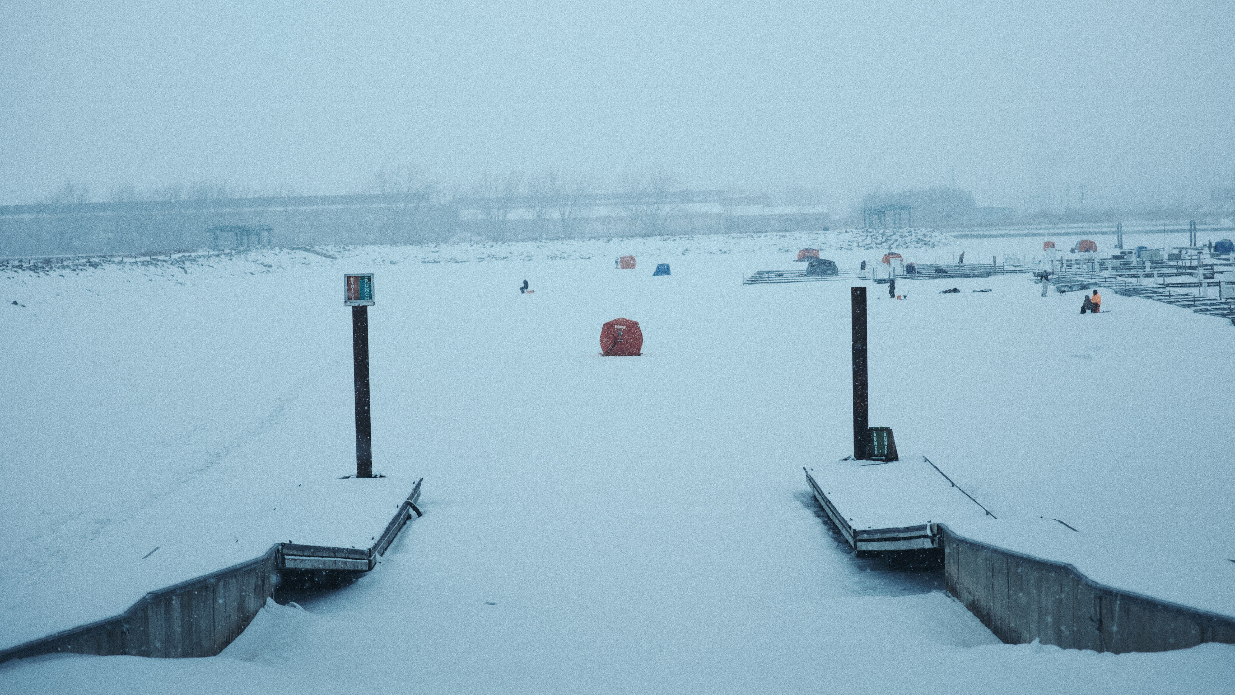 Snow-covered landscape with scattered ice fishing tents and distant trees under a pale sky.