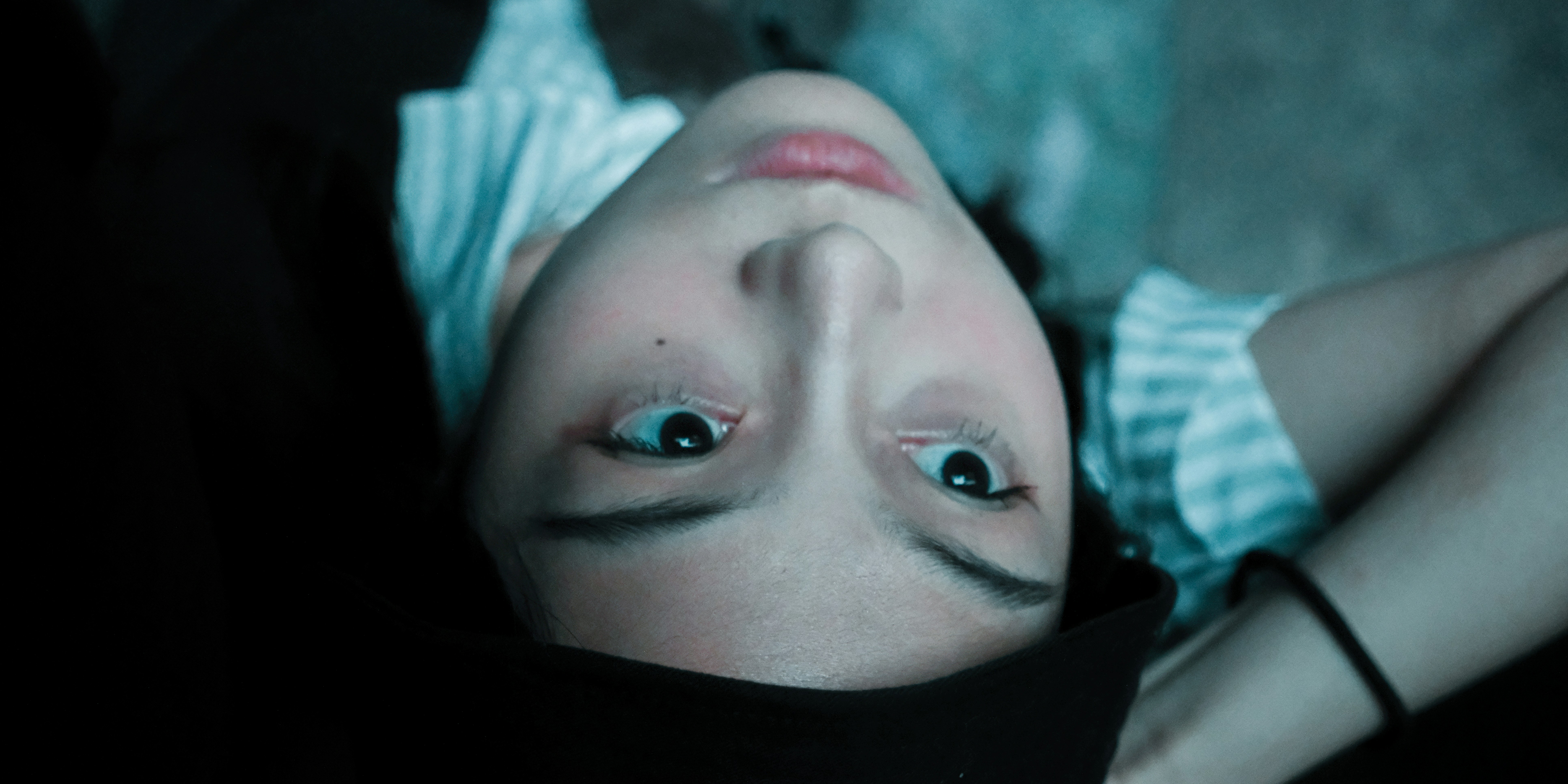 A young woman lies on her back, gazing thoughtfully upwards, with her striking blue eyes contrasting against a dark background.