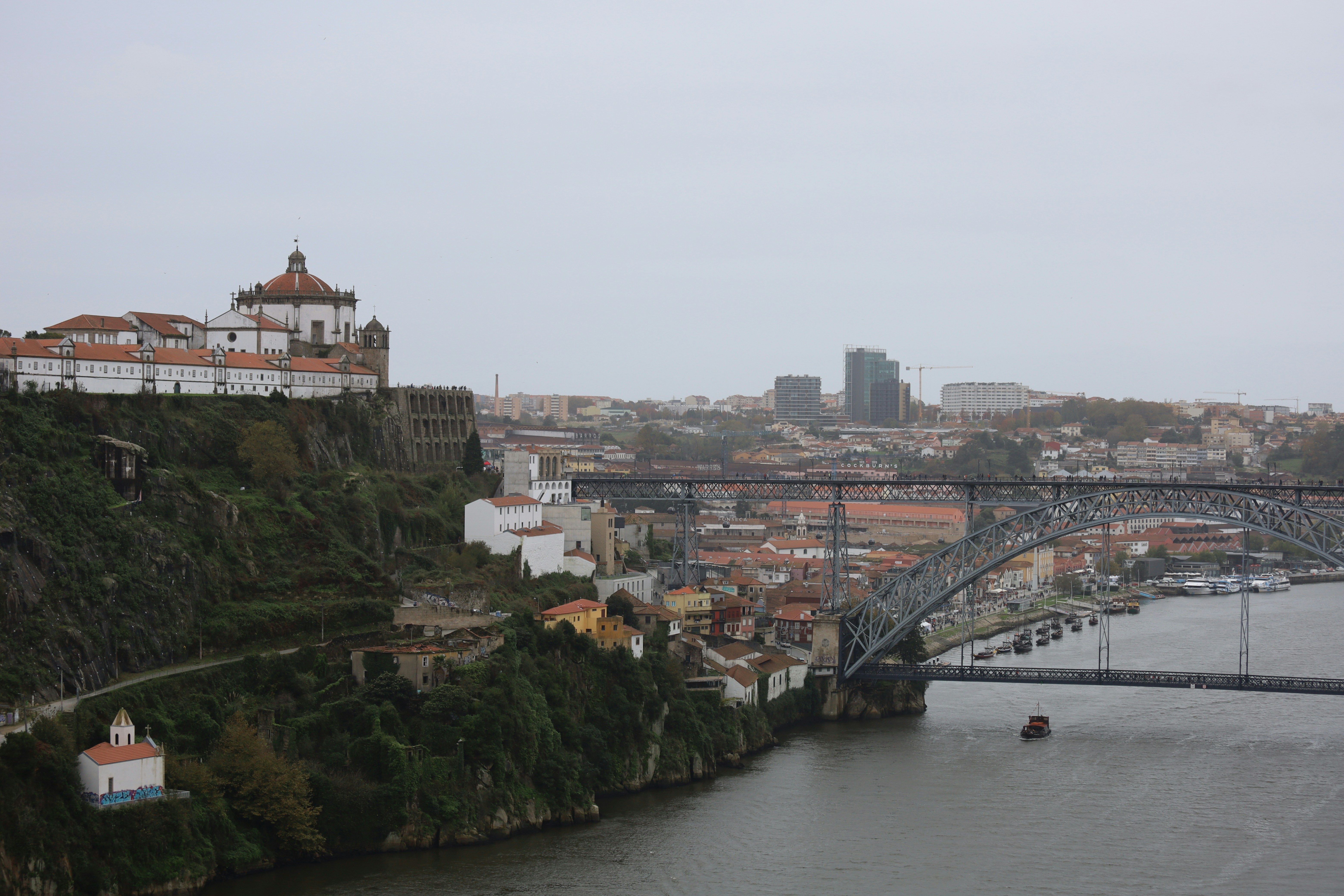 Cityscape with a historic monastery on a cliff and an arched metal bridge spanning a river.