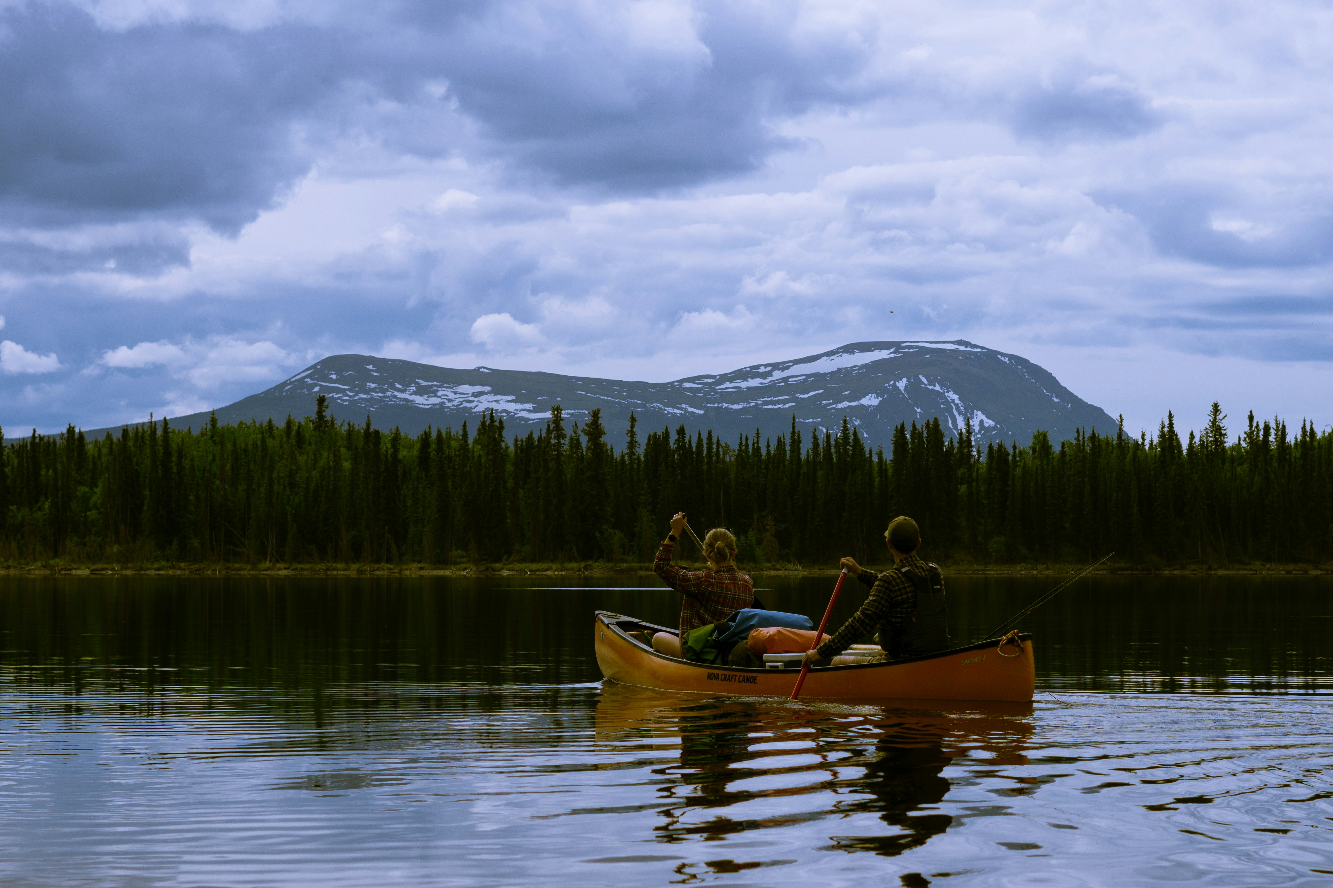Two people canoeing on a serene lake under a cloudy sky with mountains in the distance.