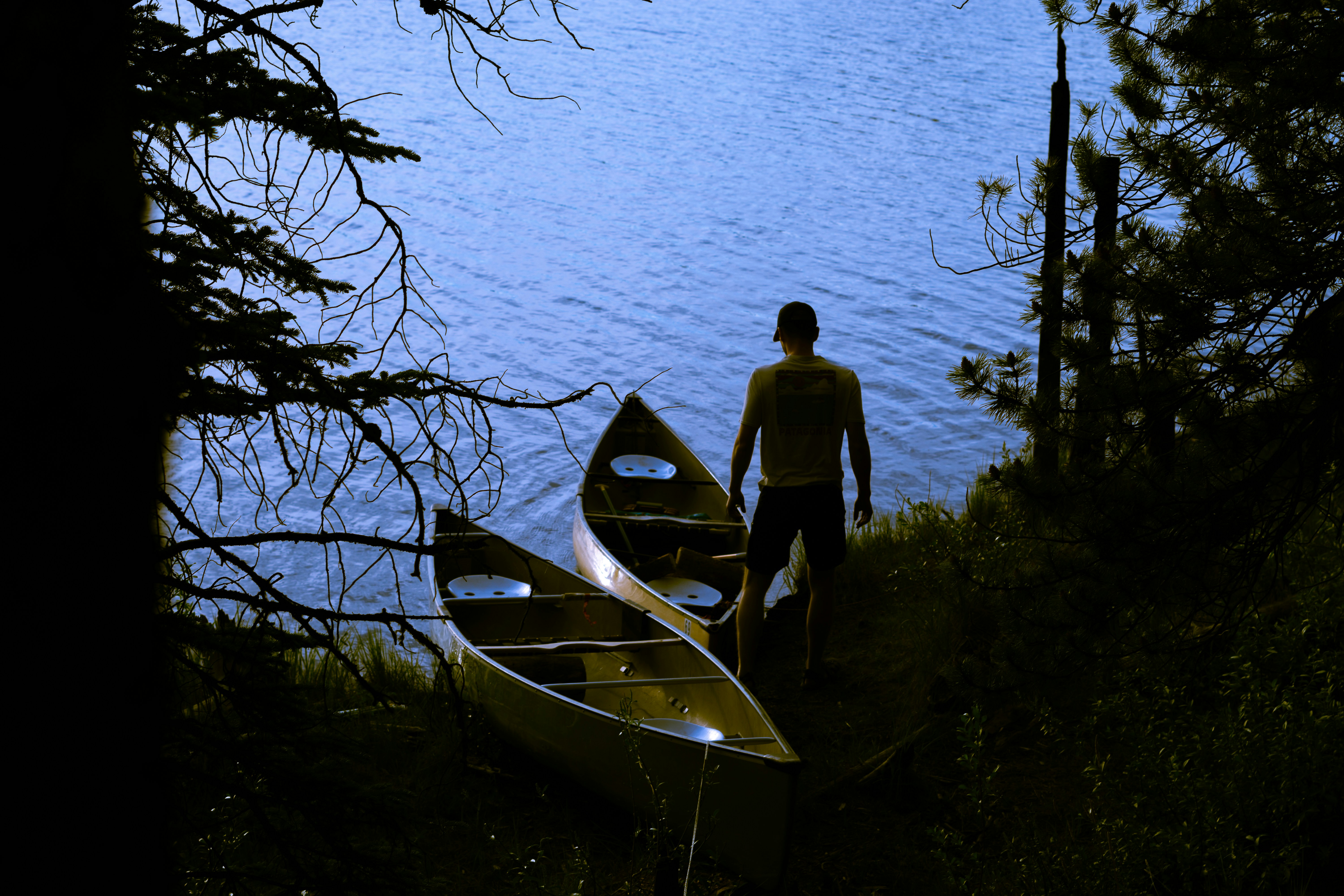 Silhouette of a person beside canoes at a wooded lakeshore under a serene blue sky.