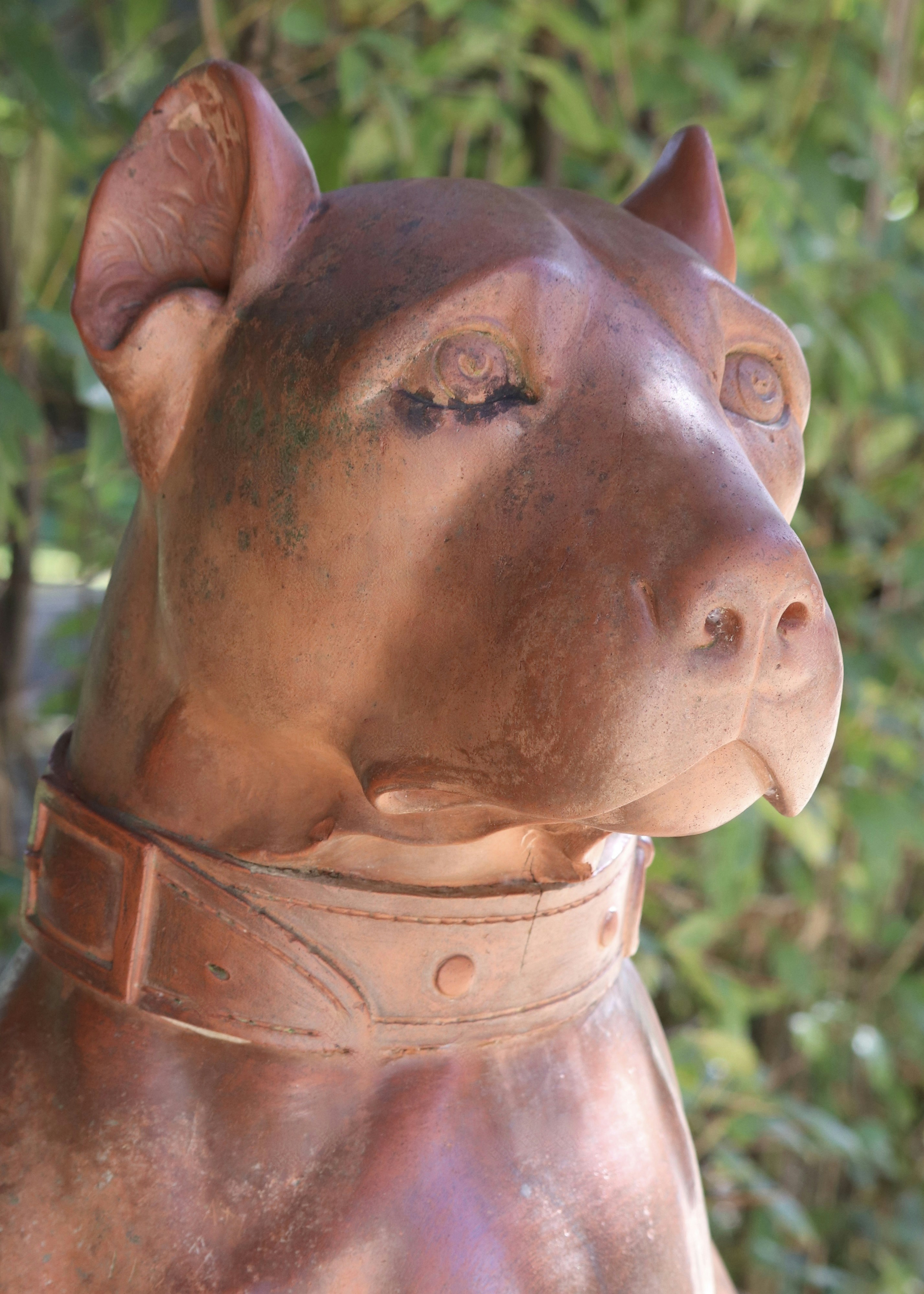 Bronze sculpture of a dog with a collar, exuding a sense of loyalty and strength, set against a lush green backdrop.