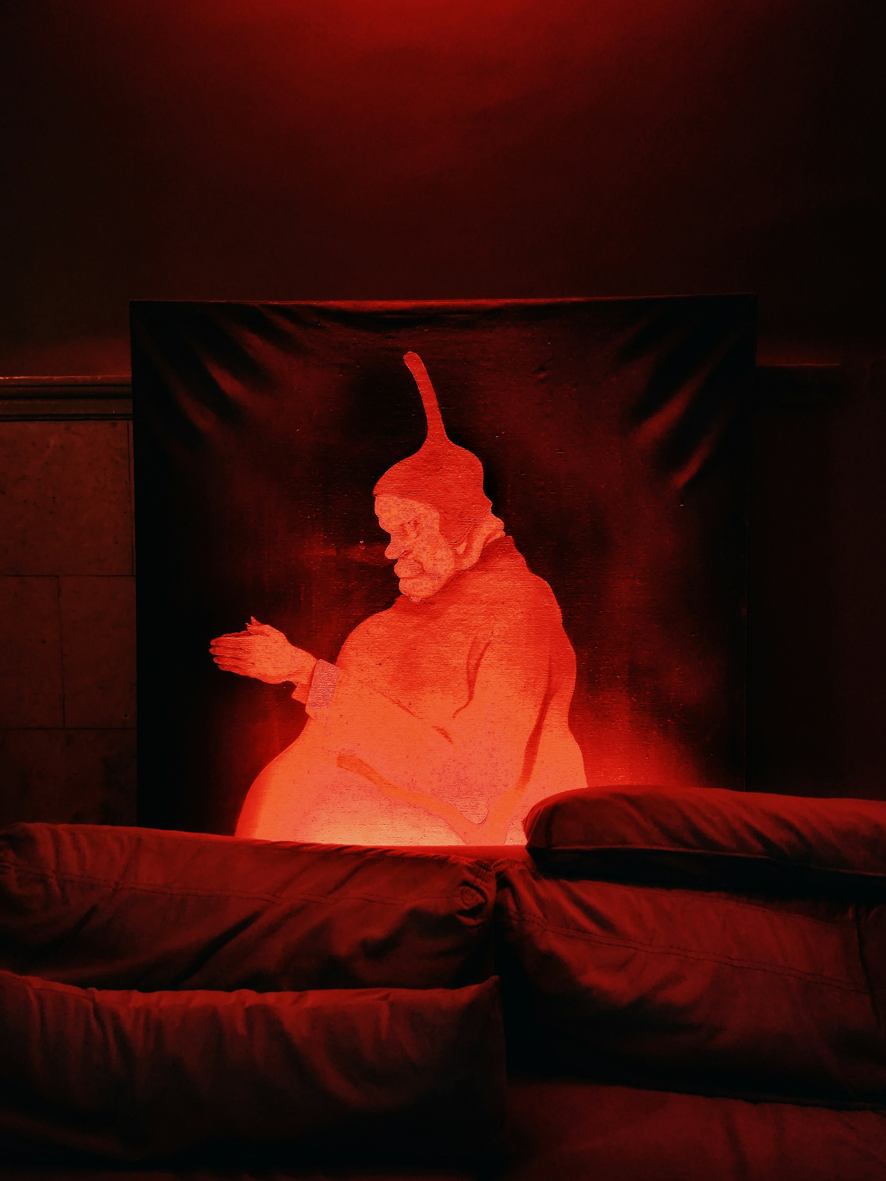 Crimson-lit painting of a seated monk-like figure hangs on a dark canvas behind a low, cushioned sofa. The deep red glow creates an intimate, contemplative mood in the room.
