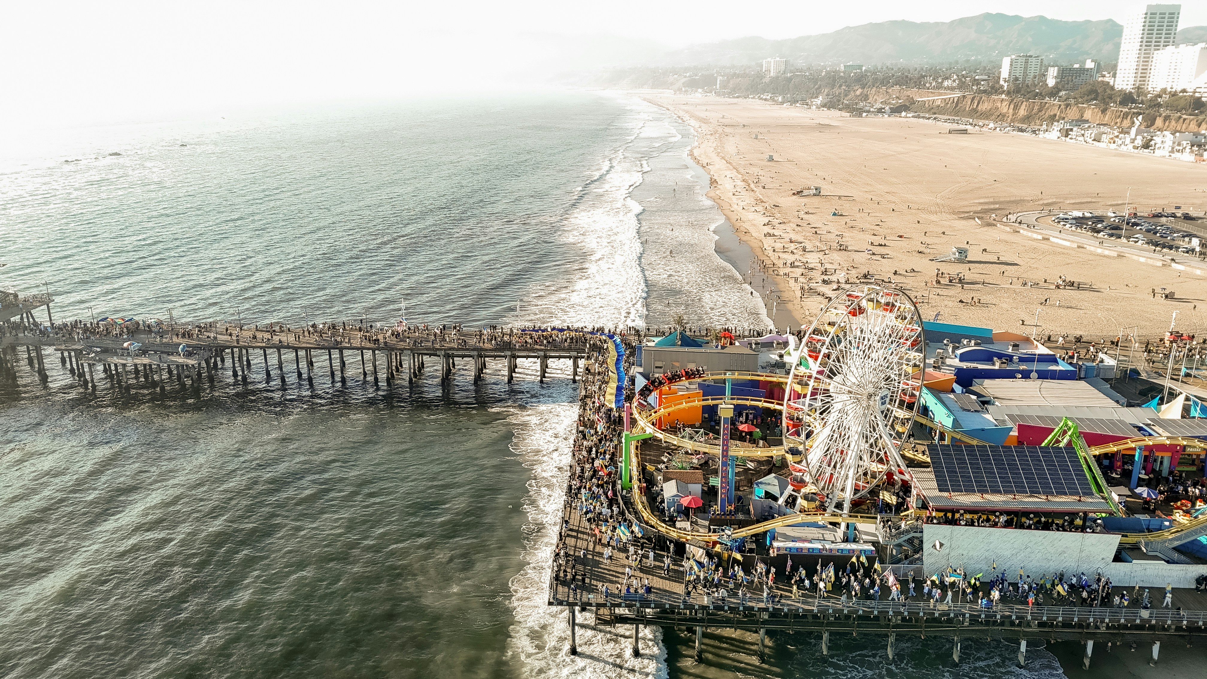 Aerial view of Santa Monica Pier adorned in blue and yellow, bustling with people near the Ferris wheel.