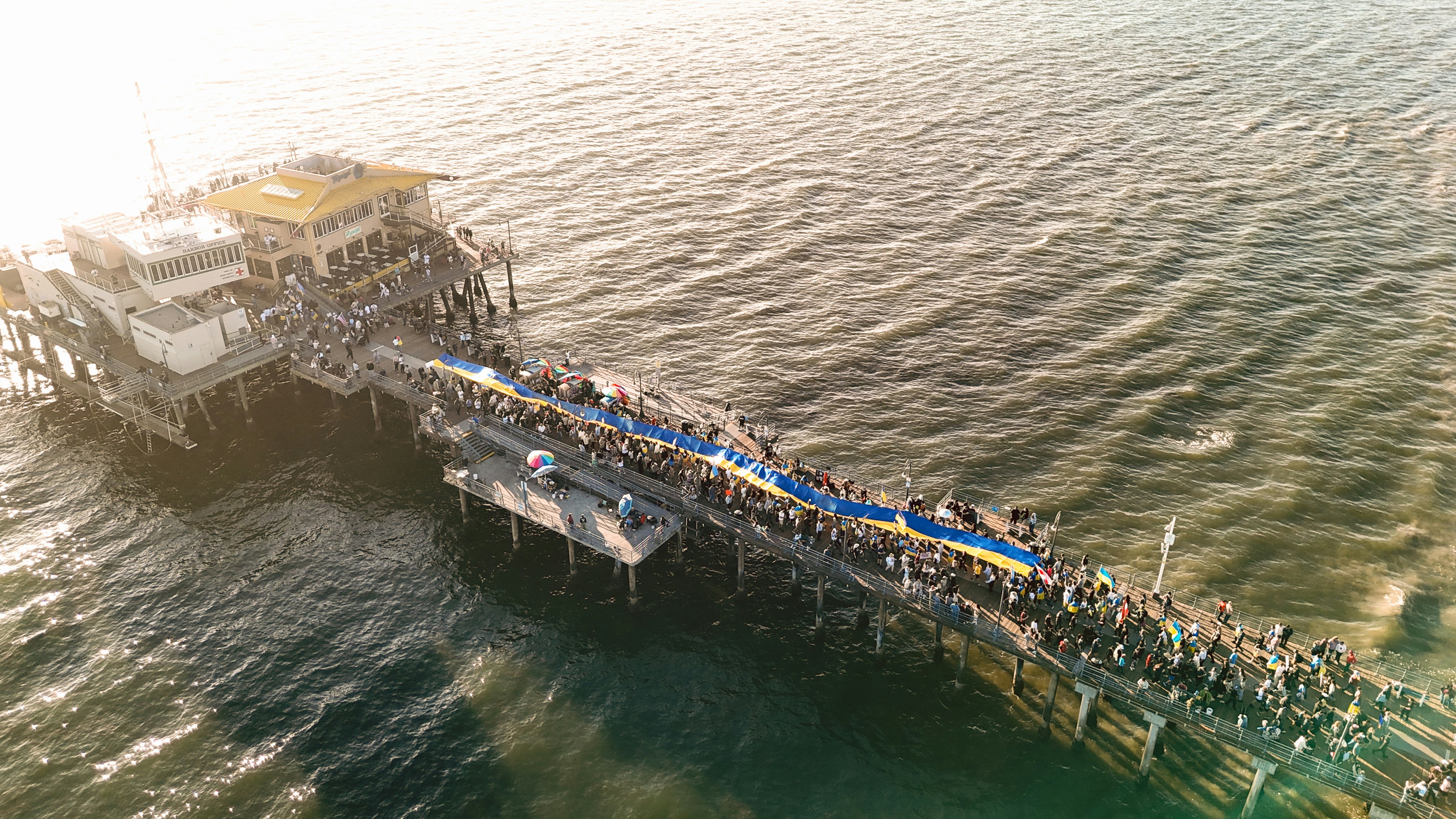 Crowded pier with a large Ukrainian flag stretching across, surrounded by shimmering ocean waves.