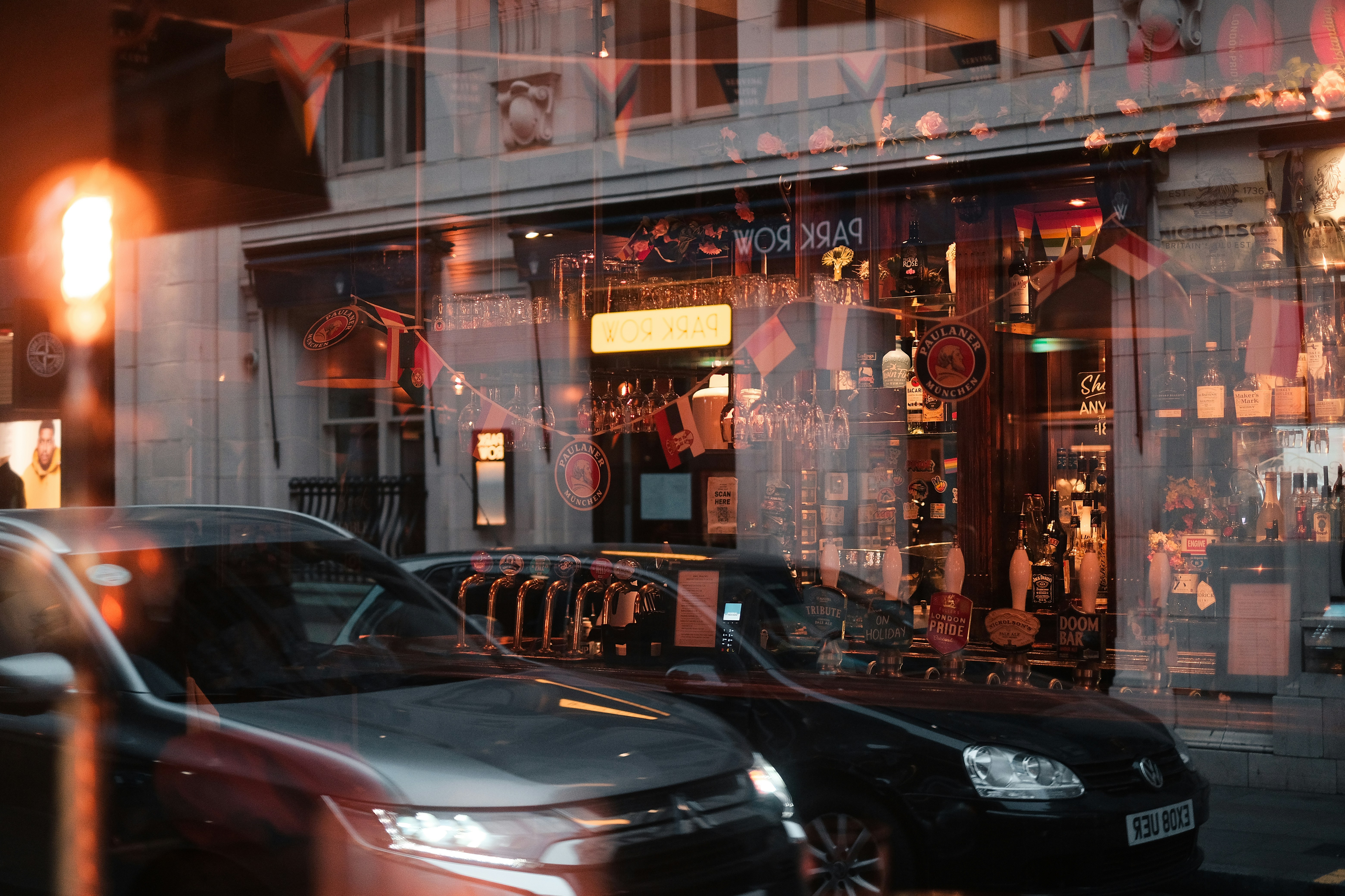 City street reflected in a pub window with warm interior lights and passing cars.