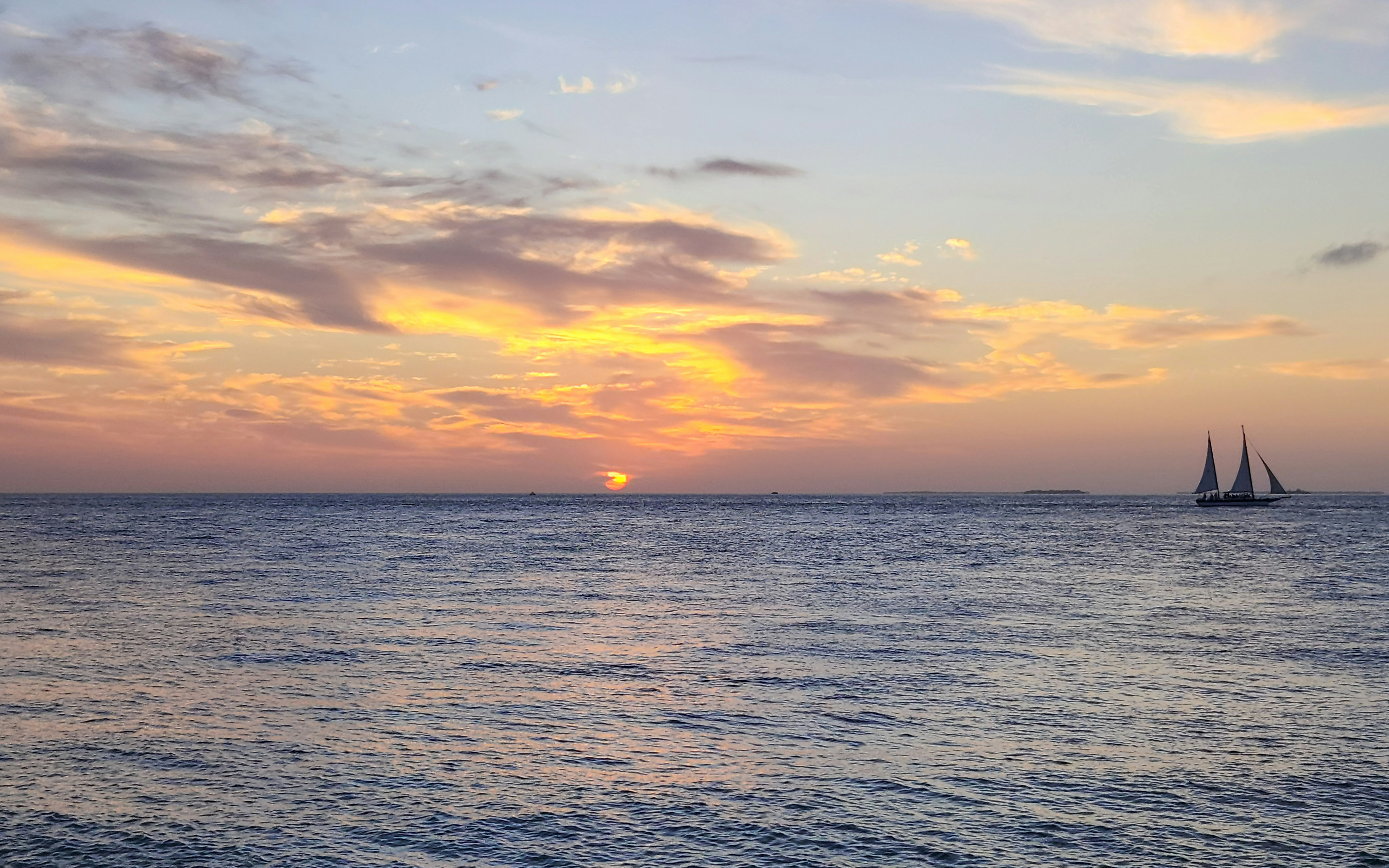 Sunset over calm ocean with a sailboat on the horizon, clouds painted in hues of orange and pink.