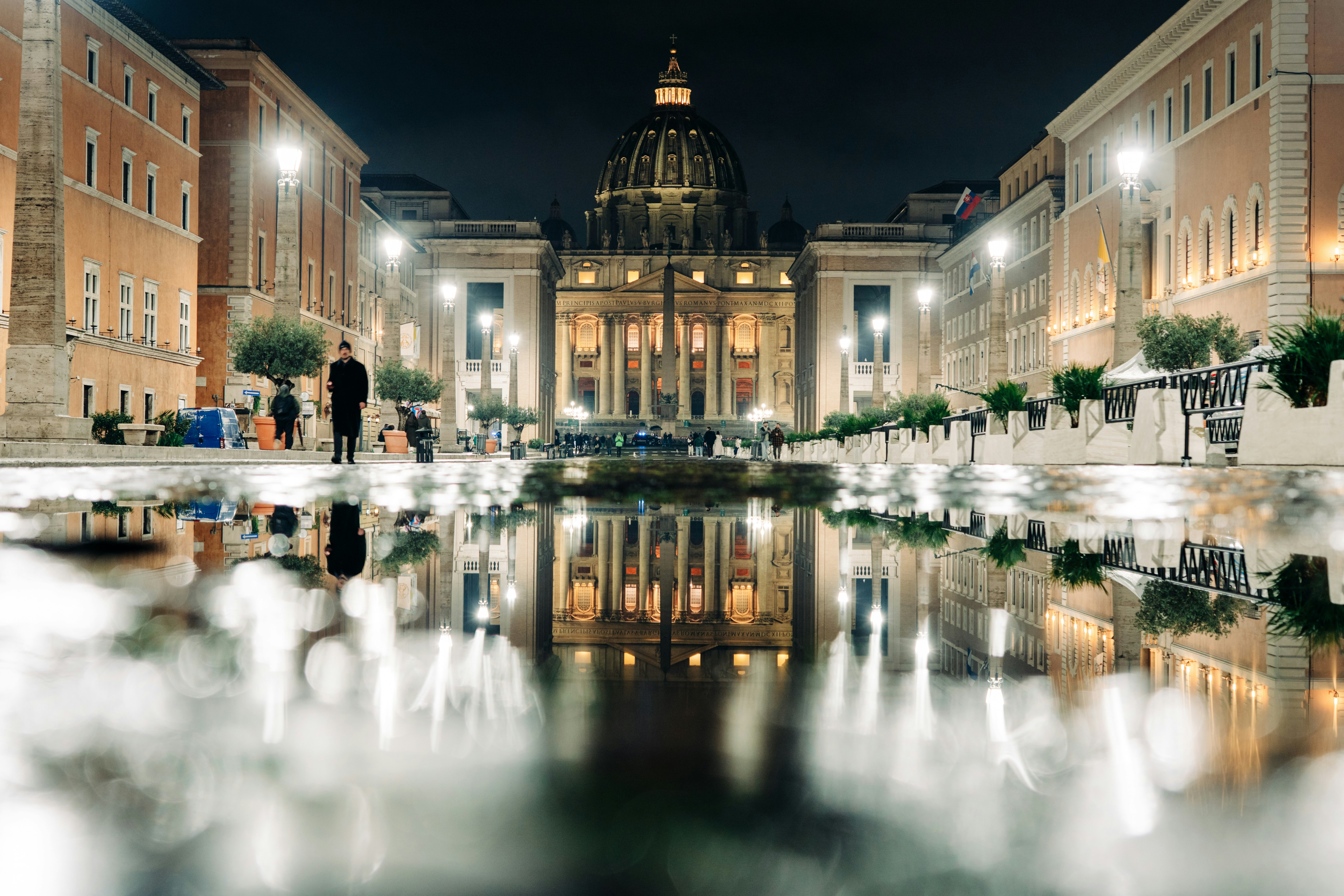 St. Peter's Basilica illuminated at night with its reflection in a puddle on the wet pavement.