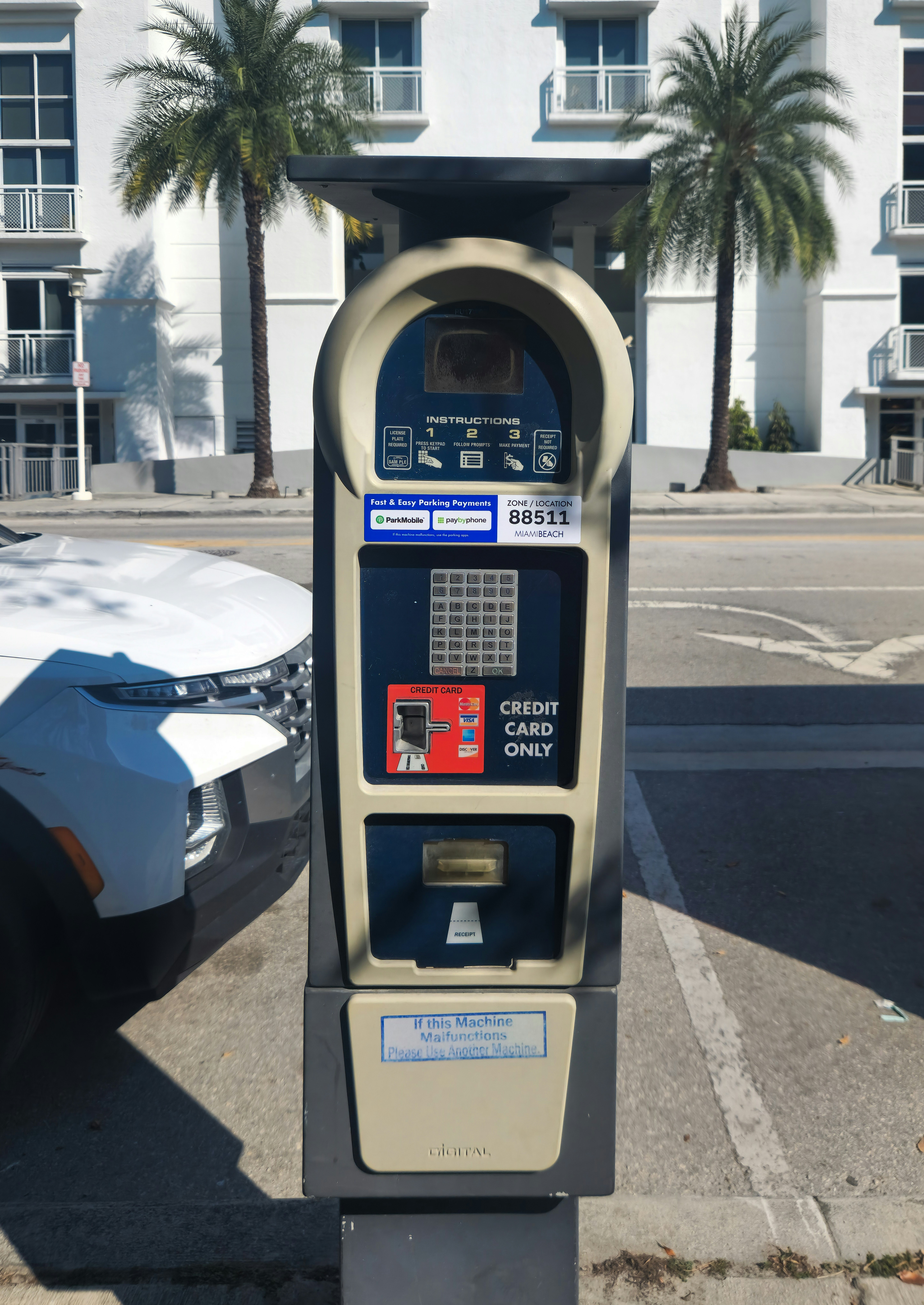 A parking meter displaying payment instructions and a credit card slot, set against a backdrop of palm trees and an urban environment.