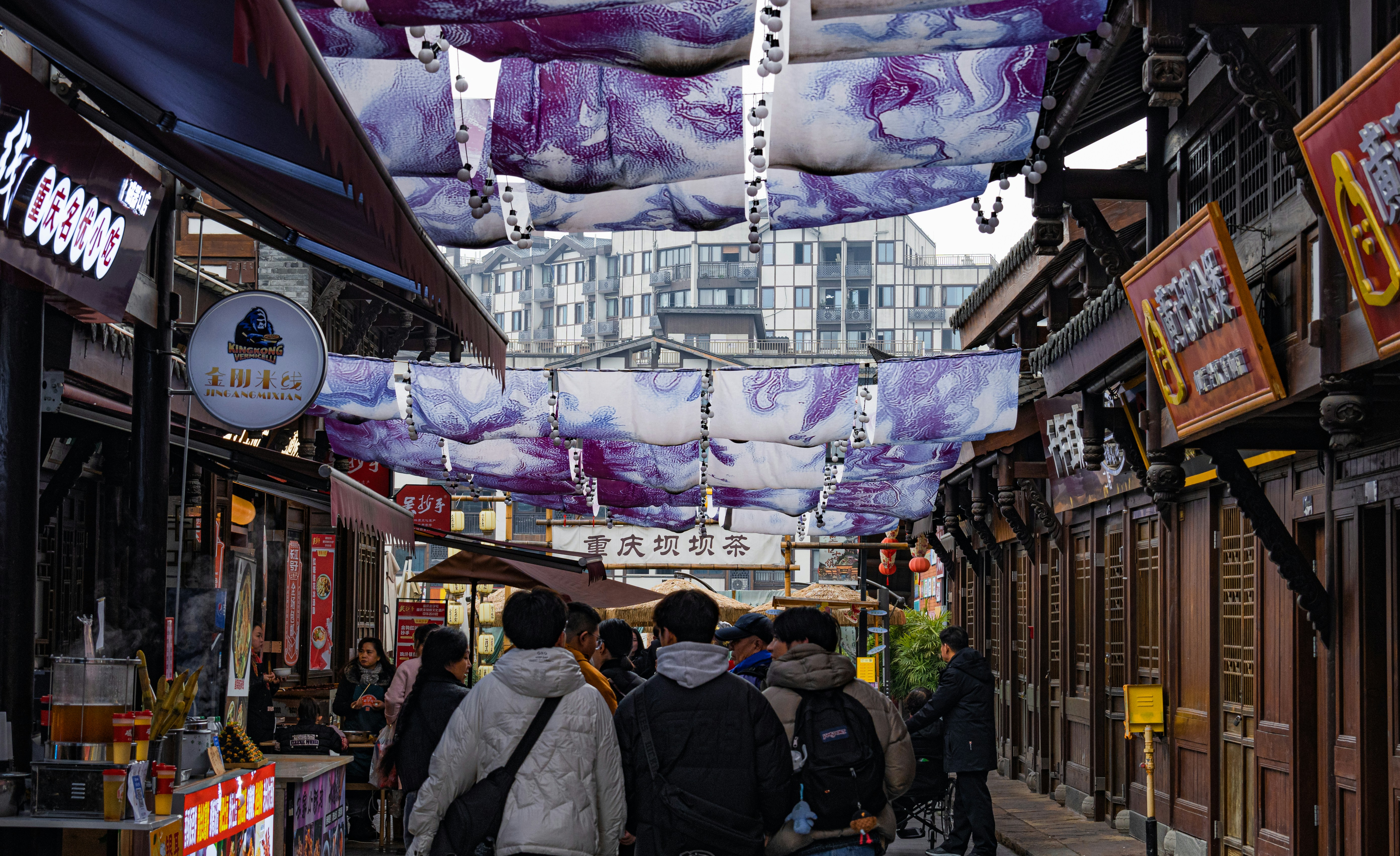 Colorful fabric canopies hang above a crowded market street lined with traditional wooden shops.