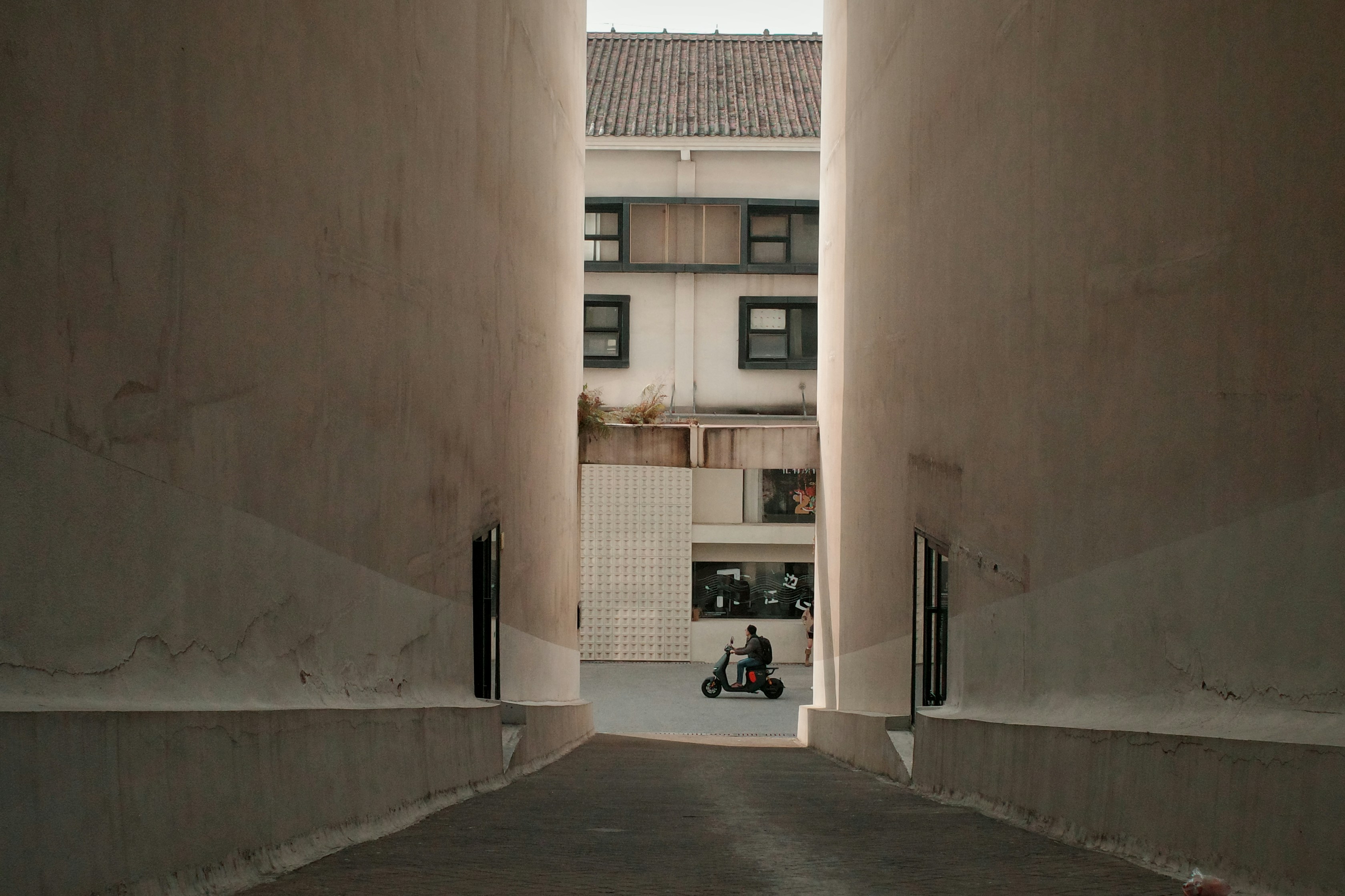 Narrow alleyway leading to a building facade with a person on a scooter in the distance.