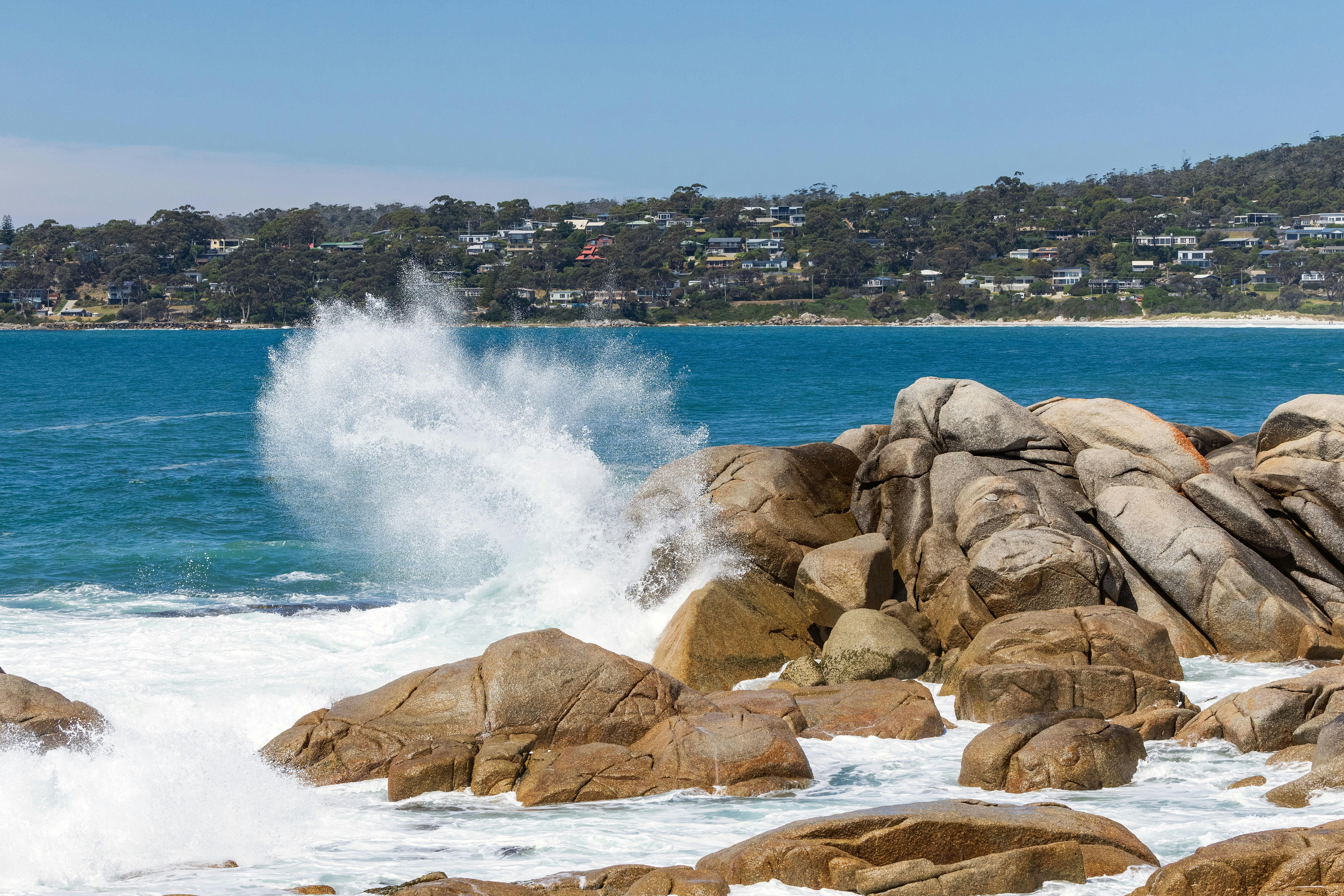 Waves crashing against rocky cliffs with coastal homes in the background under a clear blue sky.