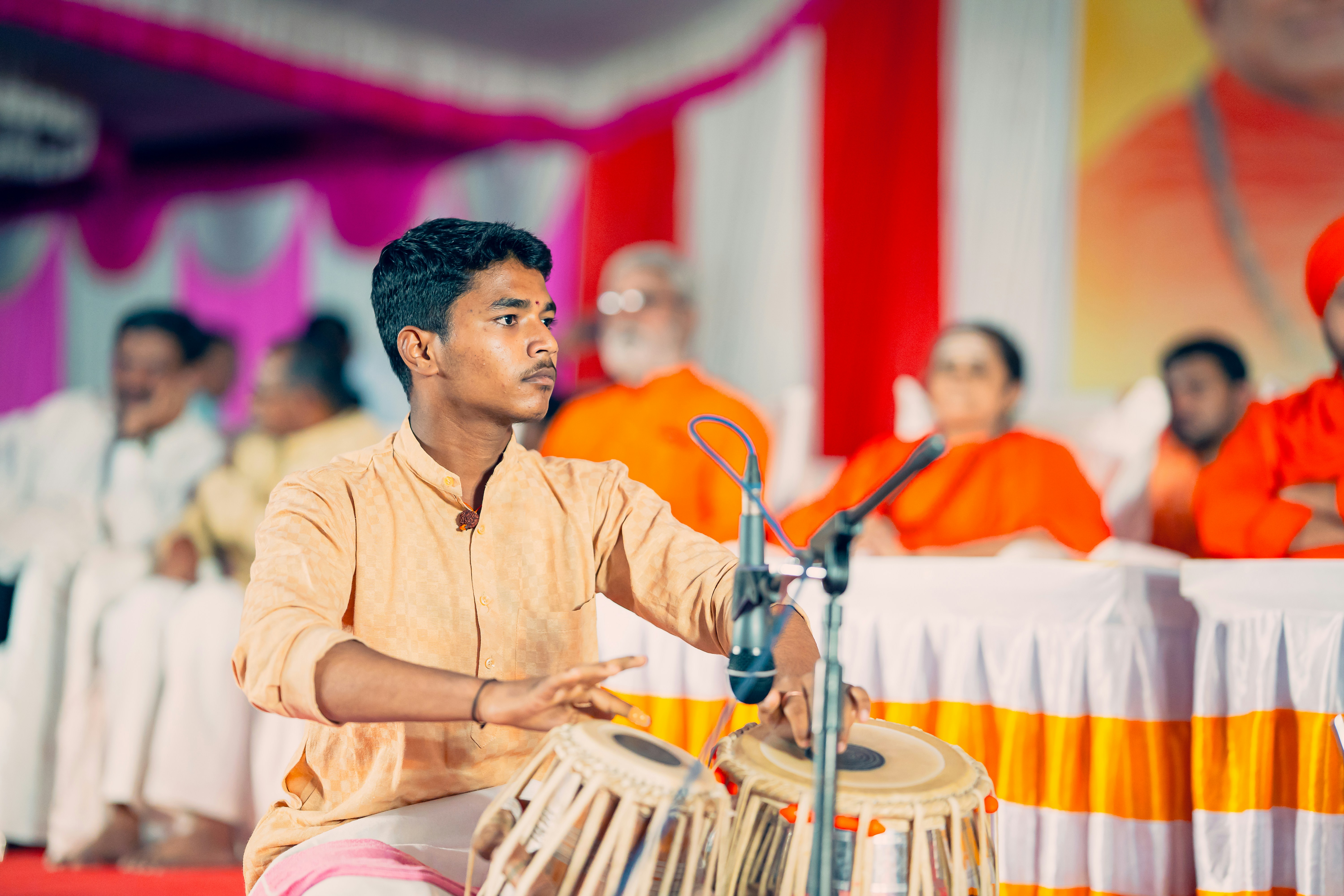 A young musician passionately playing the tabla during a cultural event, surrounded by an audience in traditional attire.