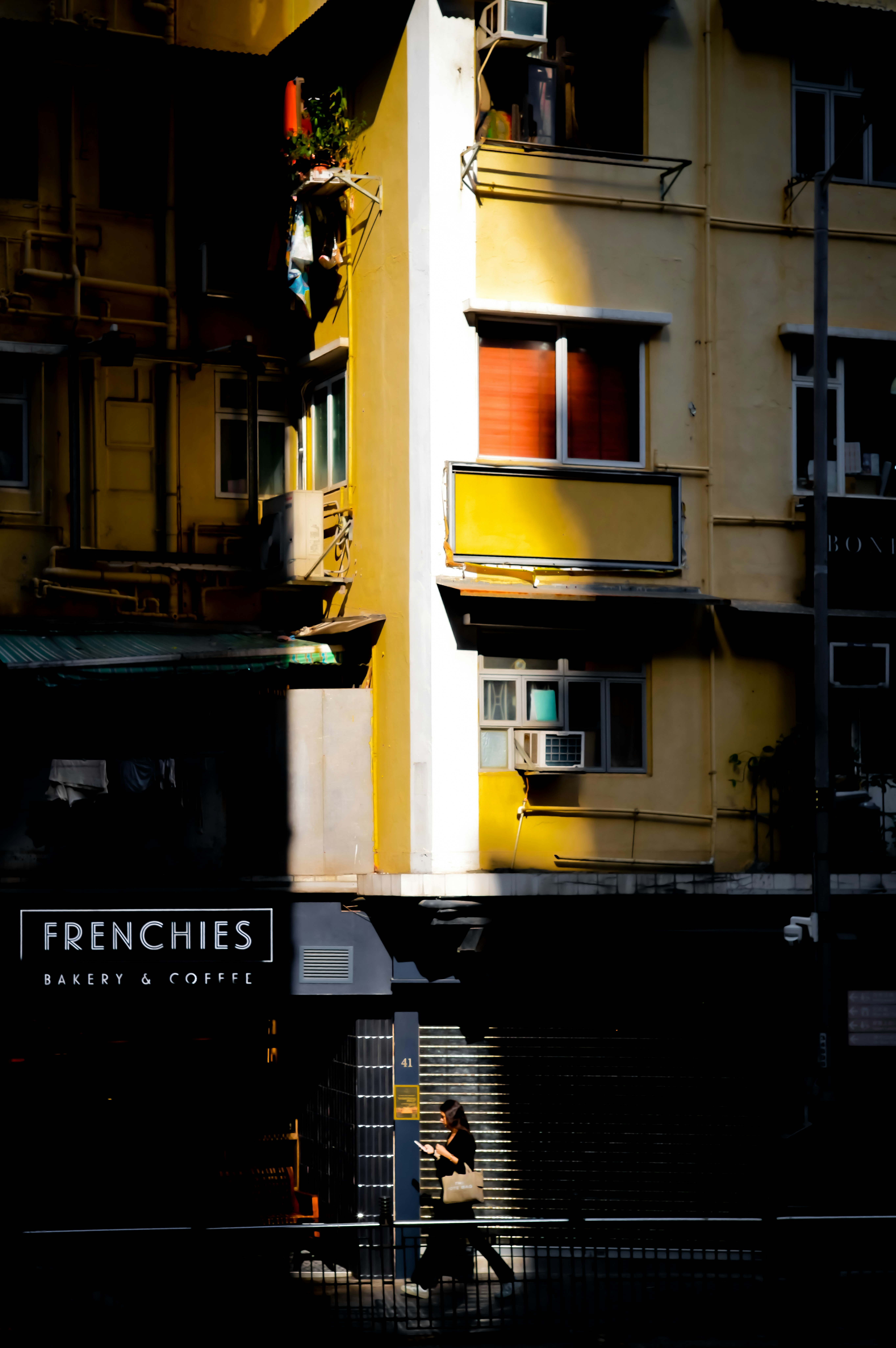 Sunlit Yellow Facade Framed by Shadow, a Pedestrian Passes by Frenchies Bakery
