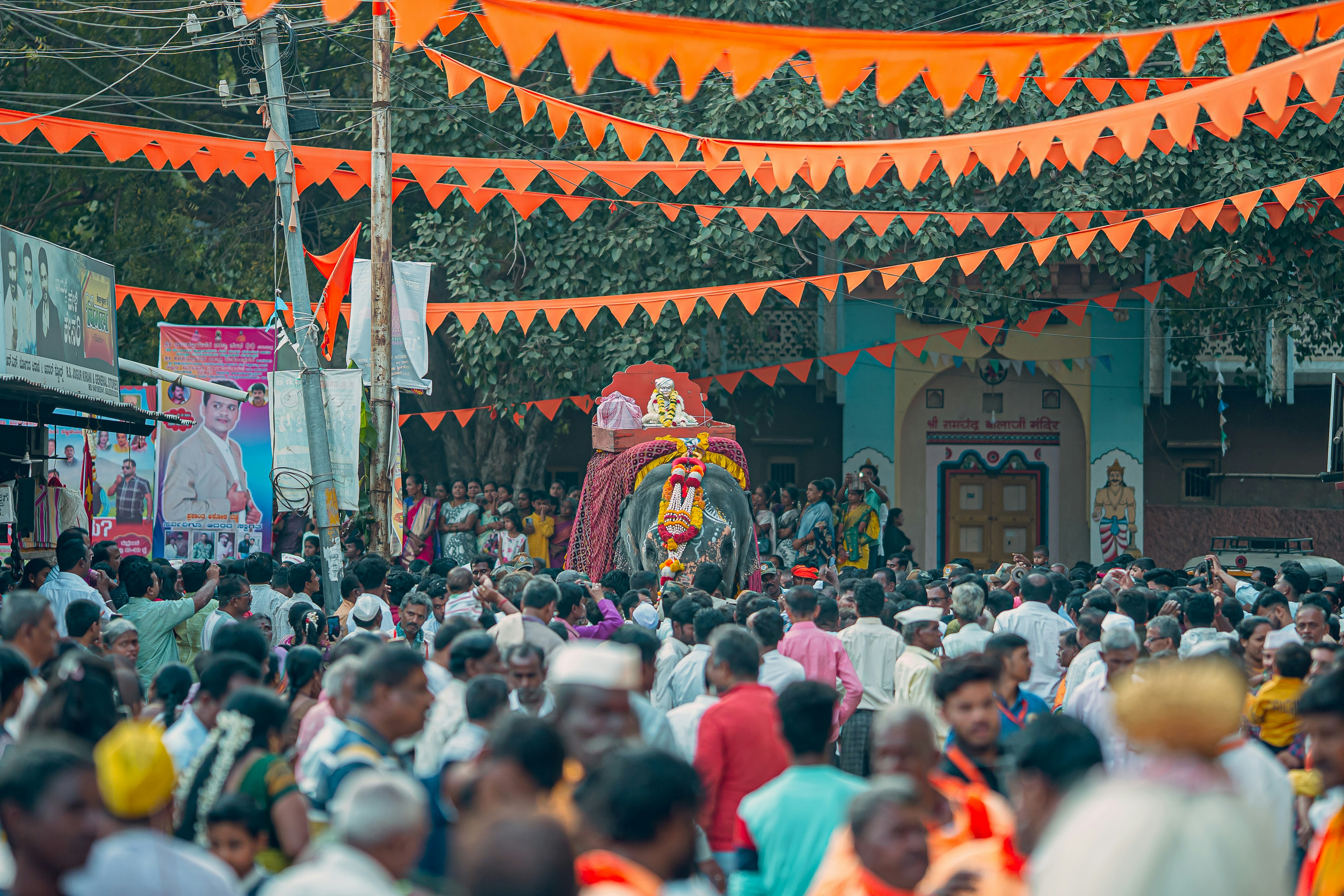 Elephant adorned with colorful decorations leads a festive crowd under orange banners.