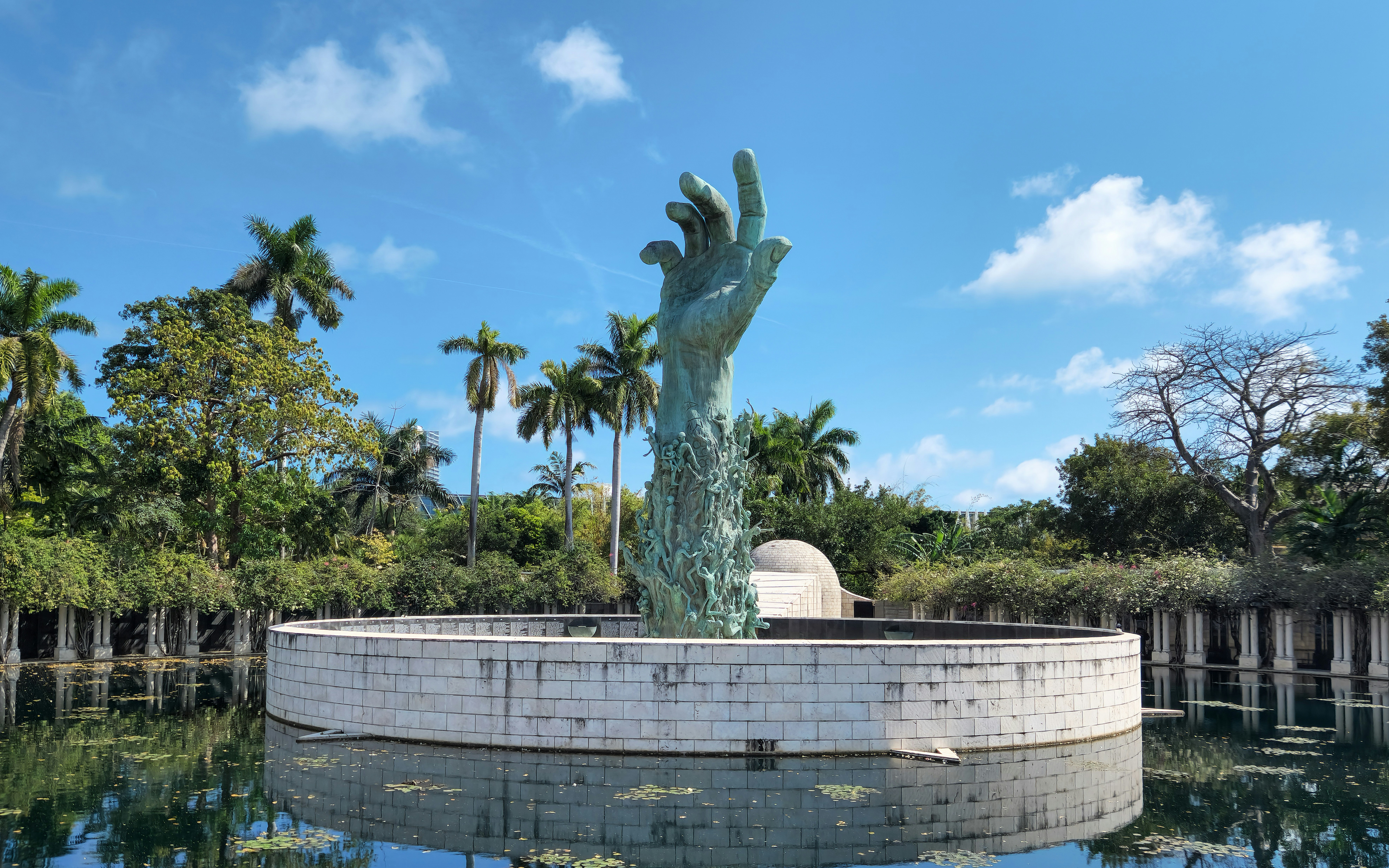 Bronze sculpture of an outstretched hand rises from a reflective pool, surrounded by lush greenery under a clear blue sky.