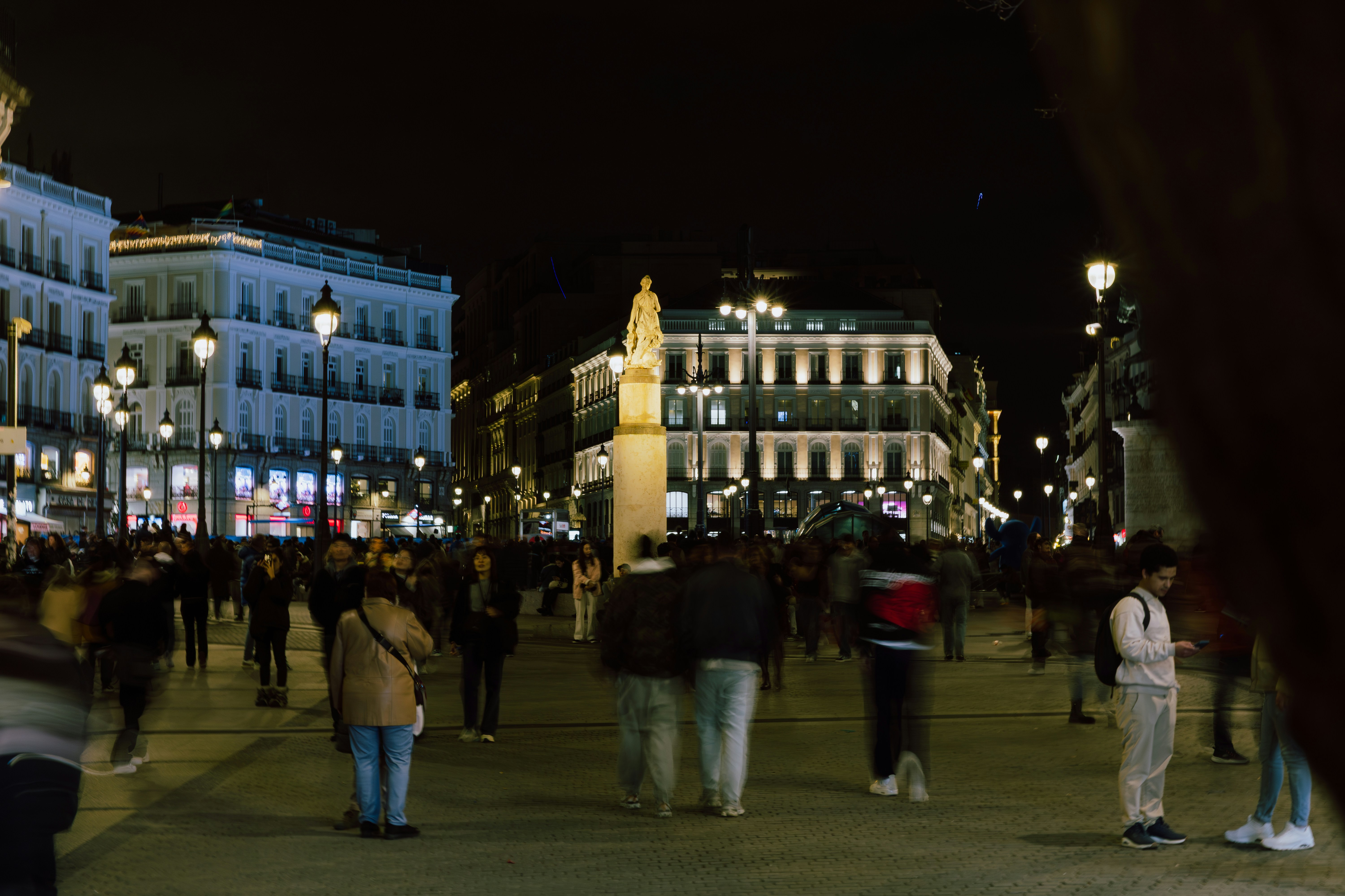 Crowds moving under lit buildings at night with a central statue illuminated.