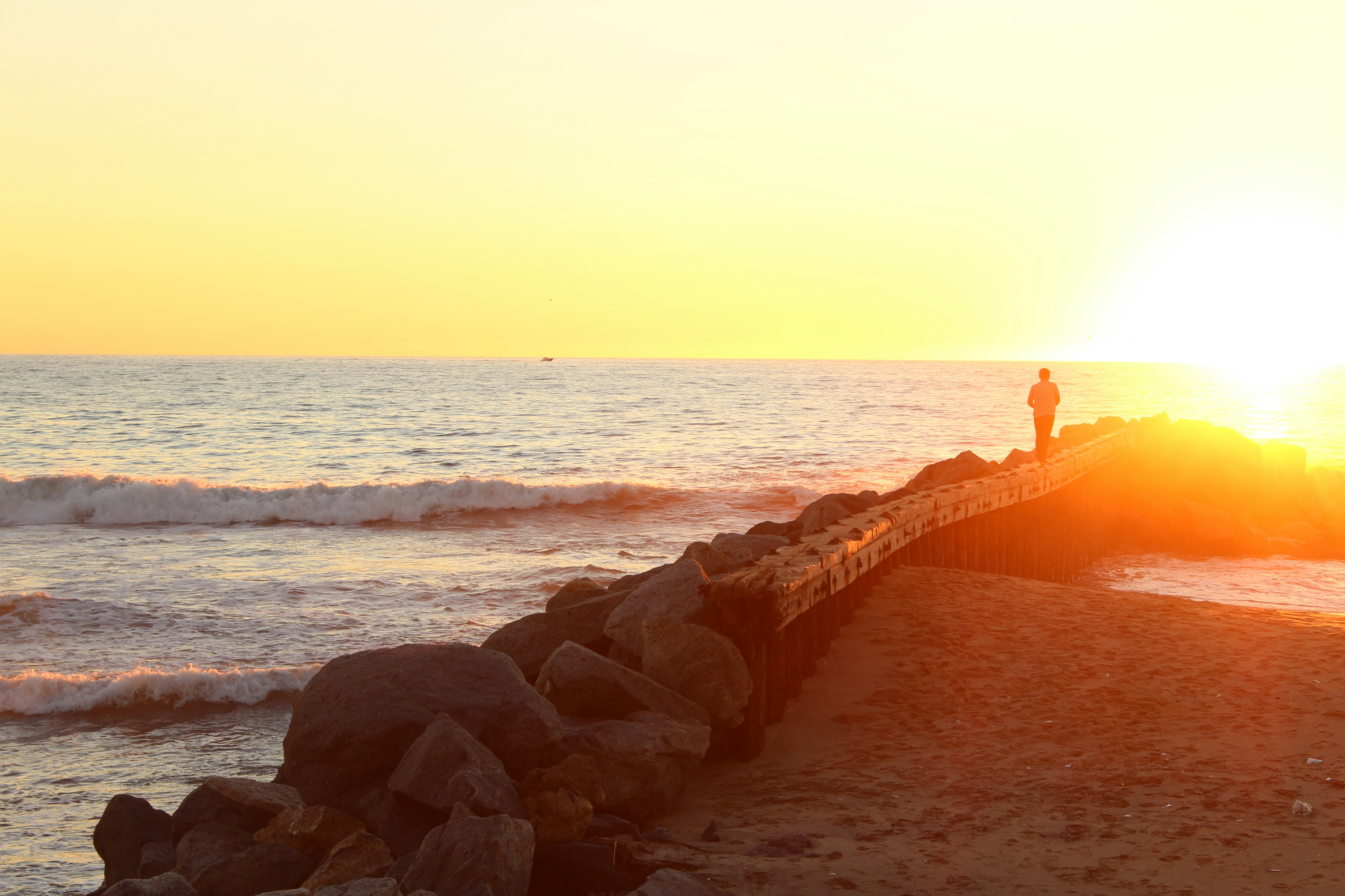 Person walking on a jetty at sunset with waves gently crashing on the shore.