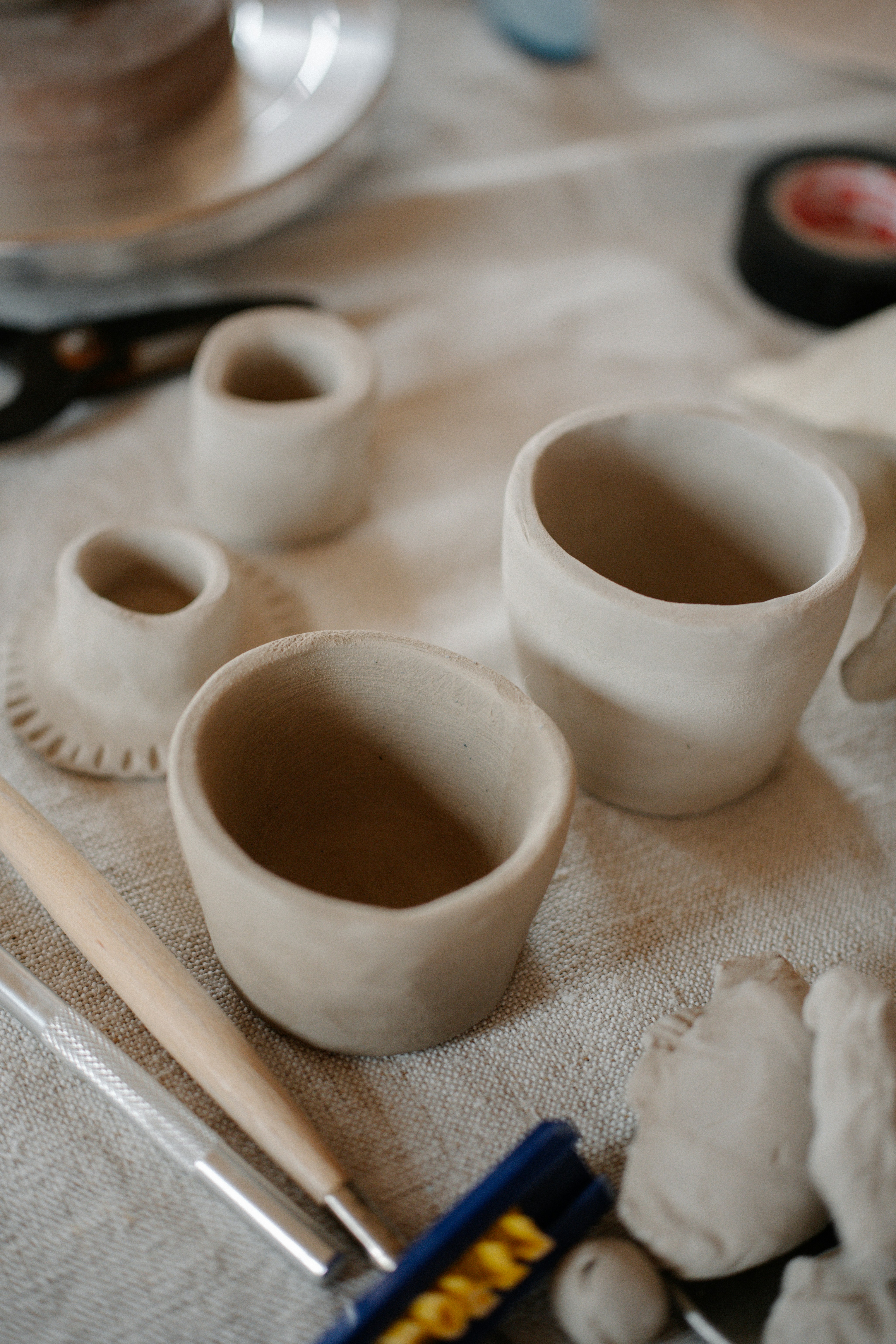 Unfired ceramic cups arranged on a textured linen surface with pottery tools nearby. A studio still-life photograph emphasizing raw clay textures and handcraft details.